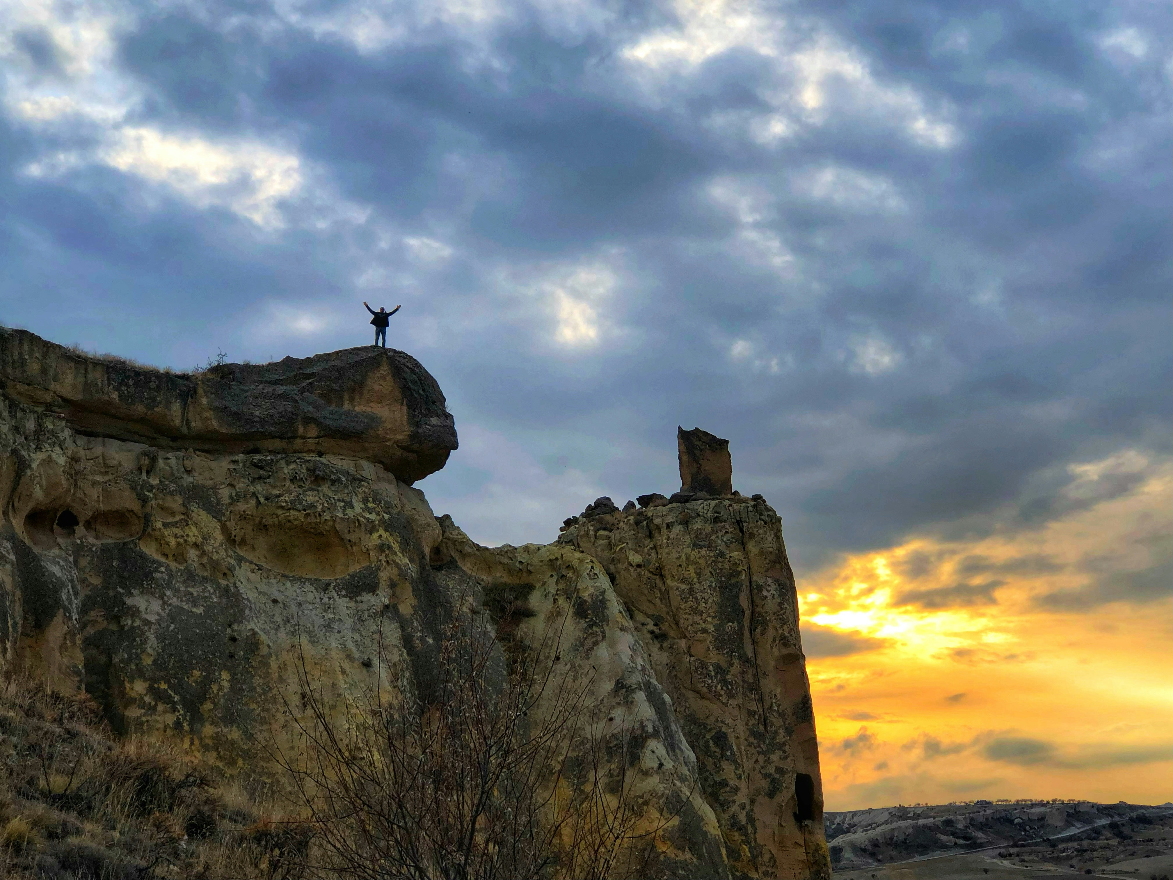 A person stands triumphantly on a rocky outcrop, silhouetted against a vibrant sunset sky filled with clouds.