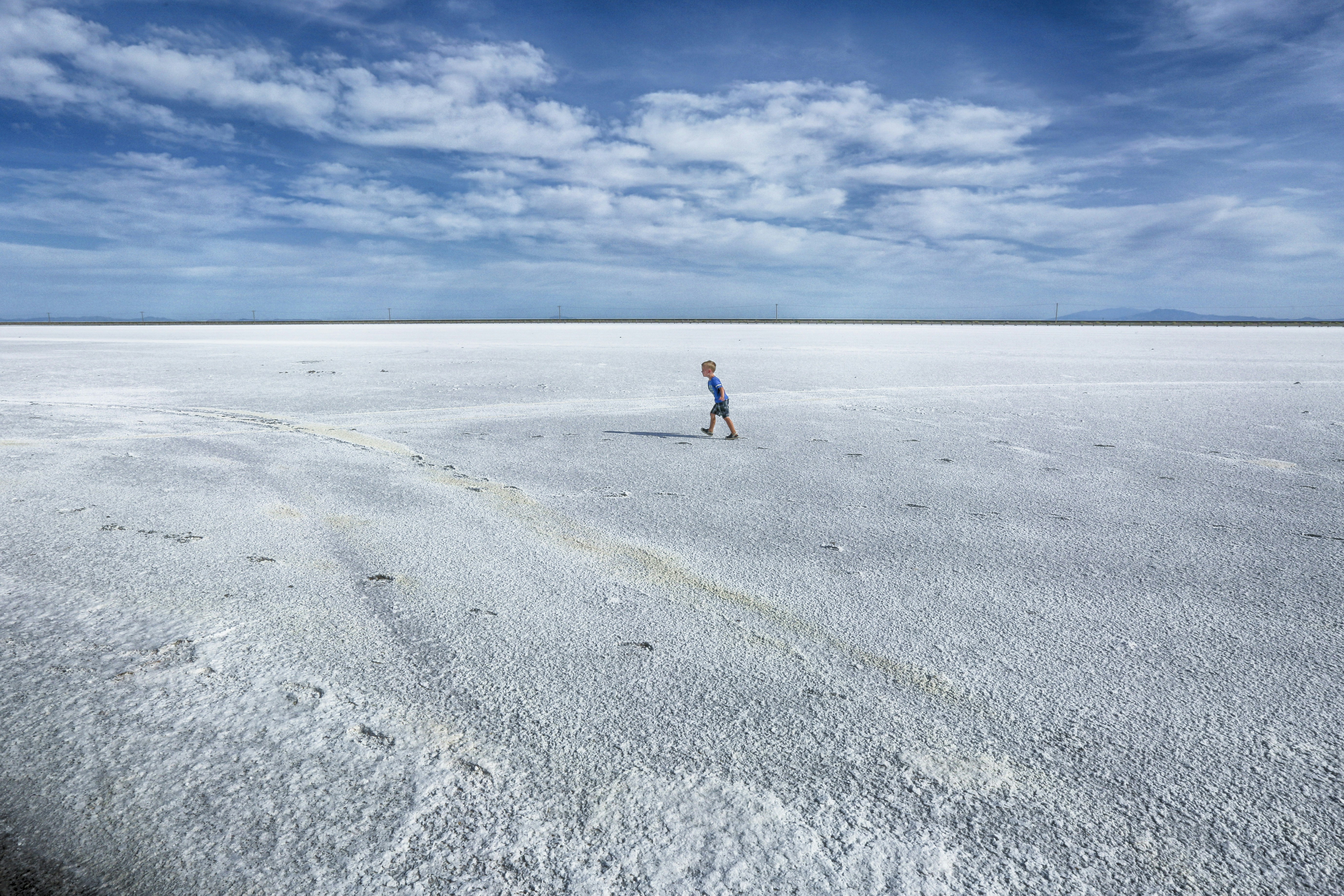 A child walks across vast, white salt flats under a blue sky with scattered clouds. The scene emphasizes the expansive, desolate beauty of the landscape.