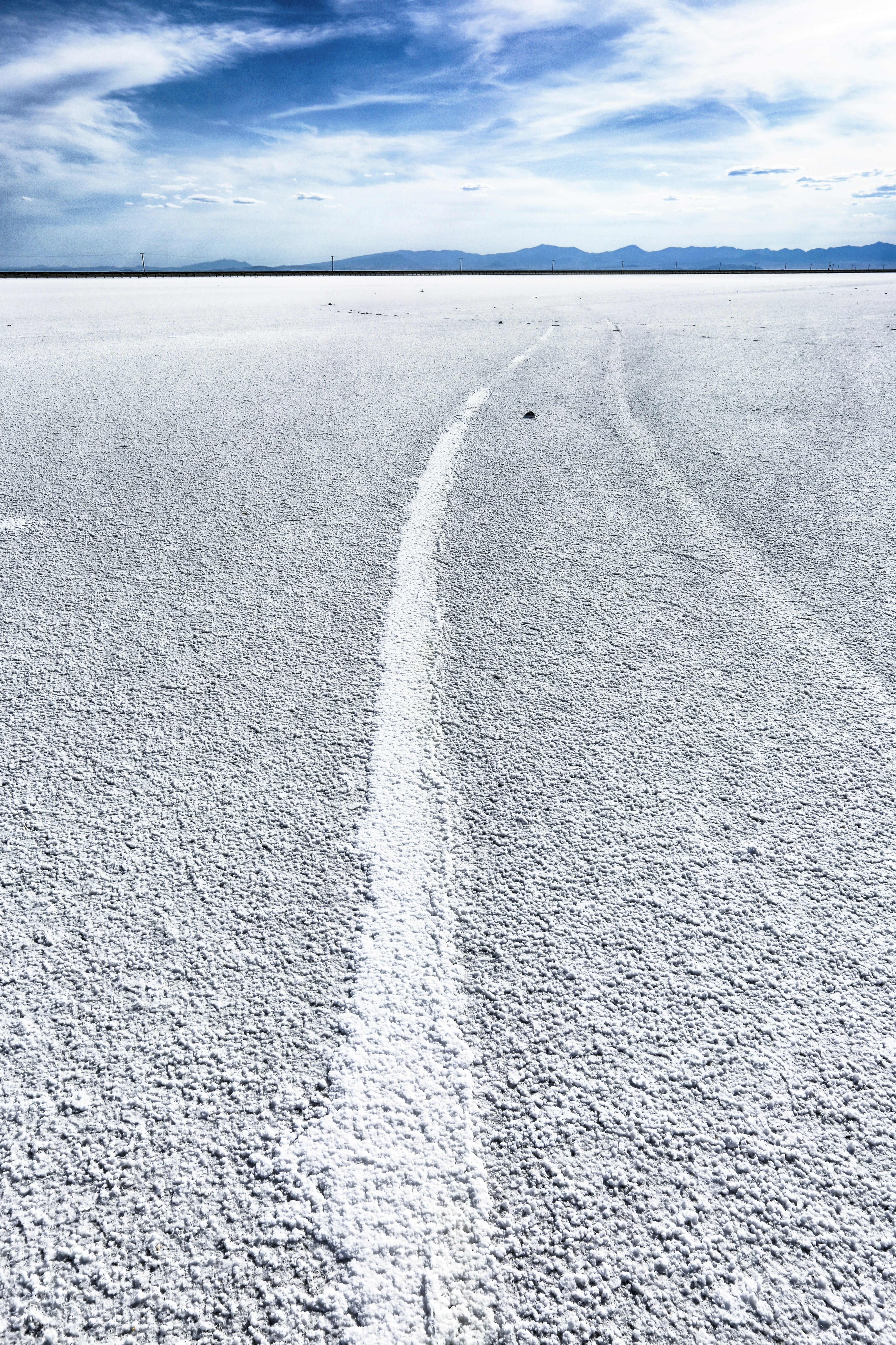 A vast expanse of salt flat with distinct tire tracks leading into the horizon under a blue sky with wispy clouds.