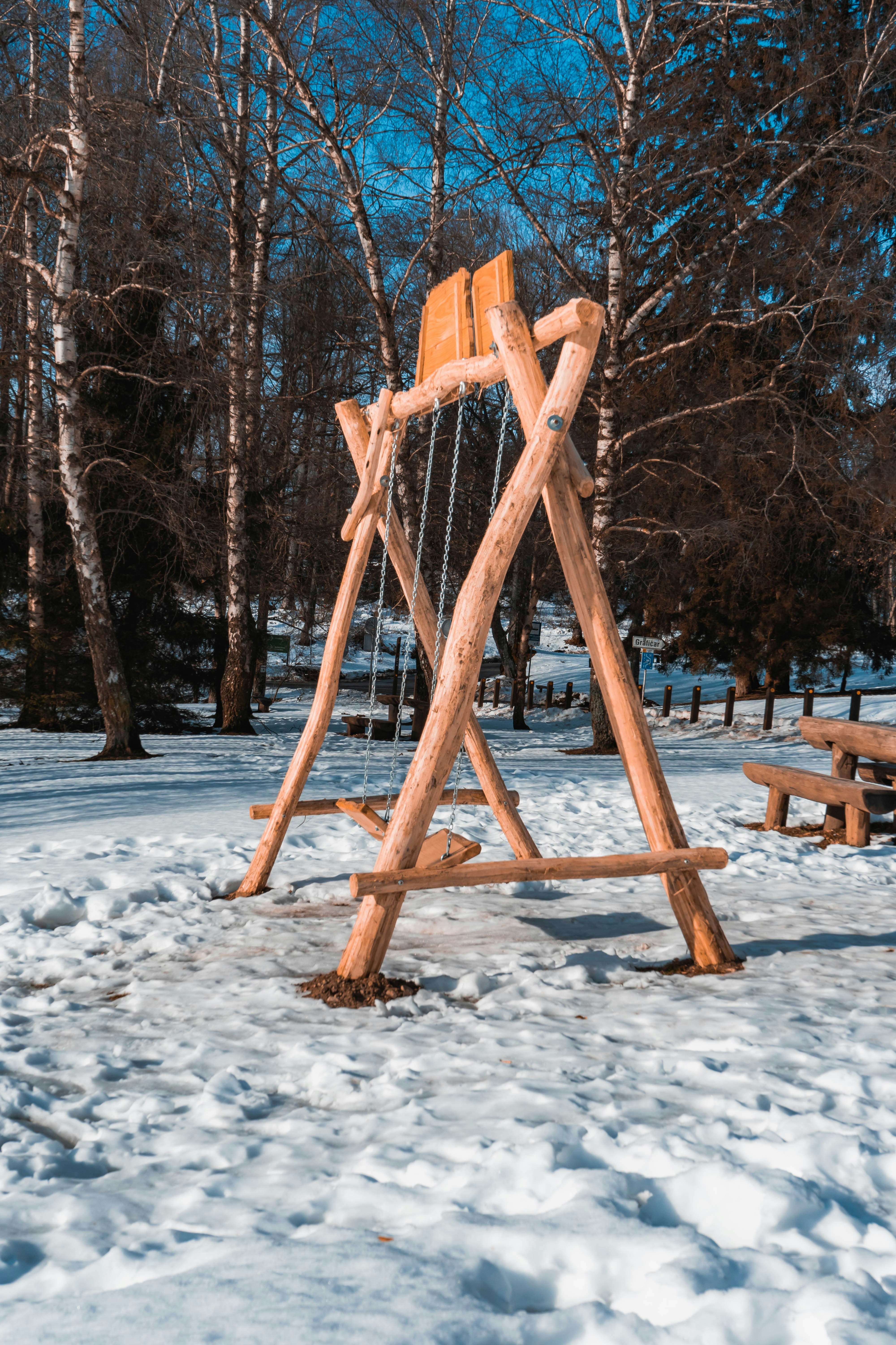 Wooden swing set stands alone in a snow-covered playground surrounded by bare trees. The scene captures the tranquility of winter's chill.