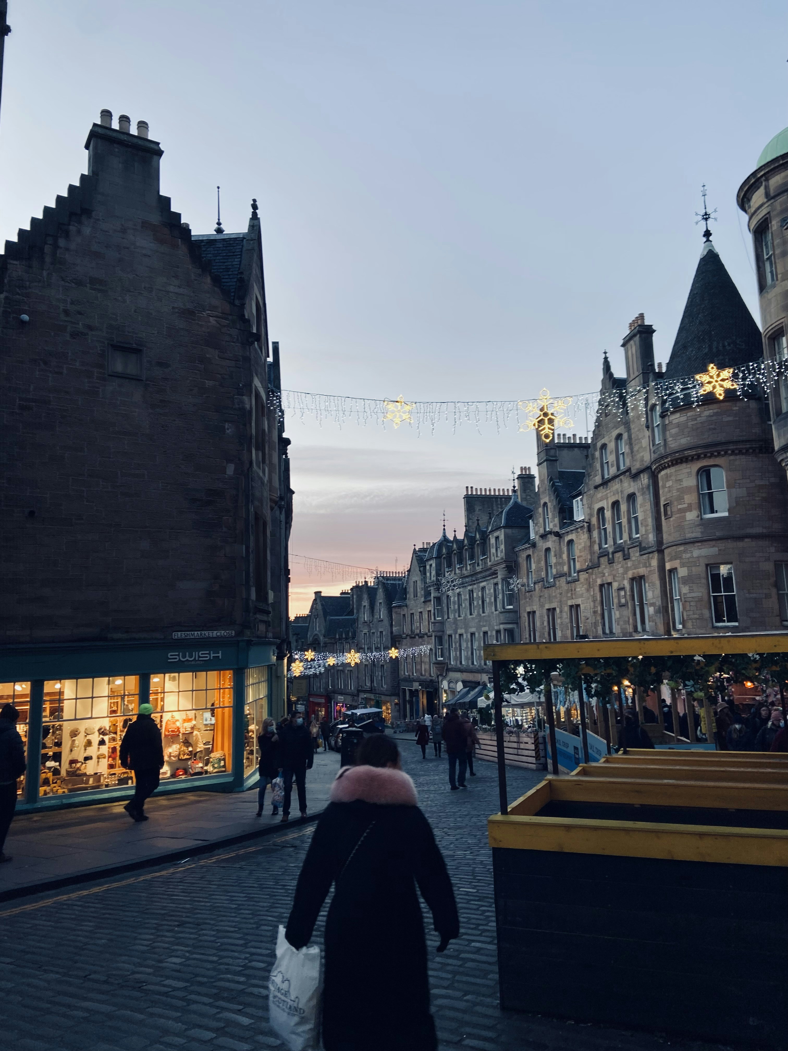 Charming street adorned with festive lights, leading into a bustling market area during twilight. The warm glow from shop windows invites passersby to explore.