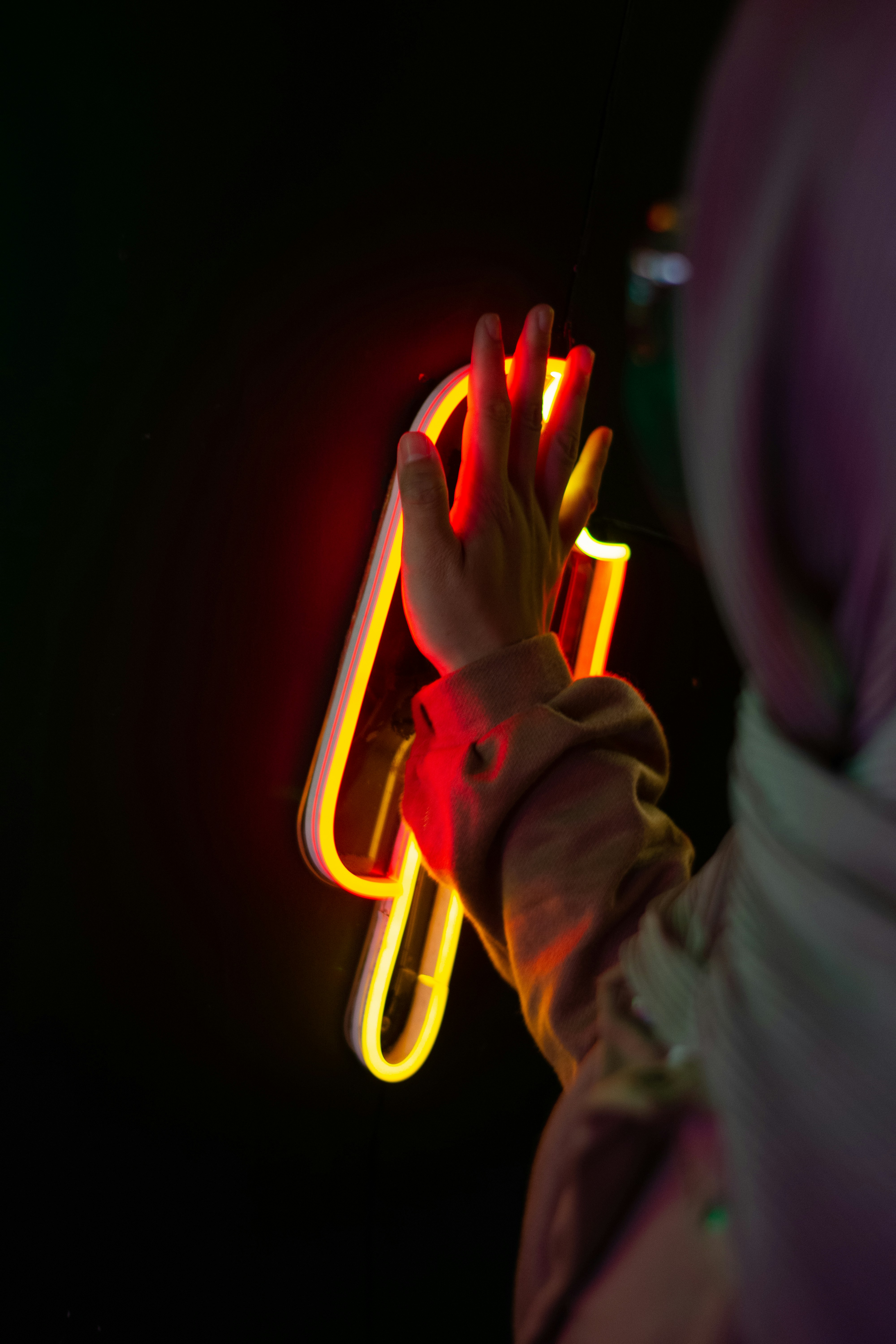 A hand reaches towards a glowing neon sign, creating a vibrant contrast against a dark background. The interplay of colors emphasizes the moment of interaction.