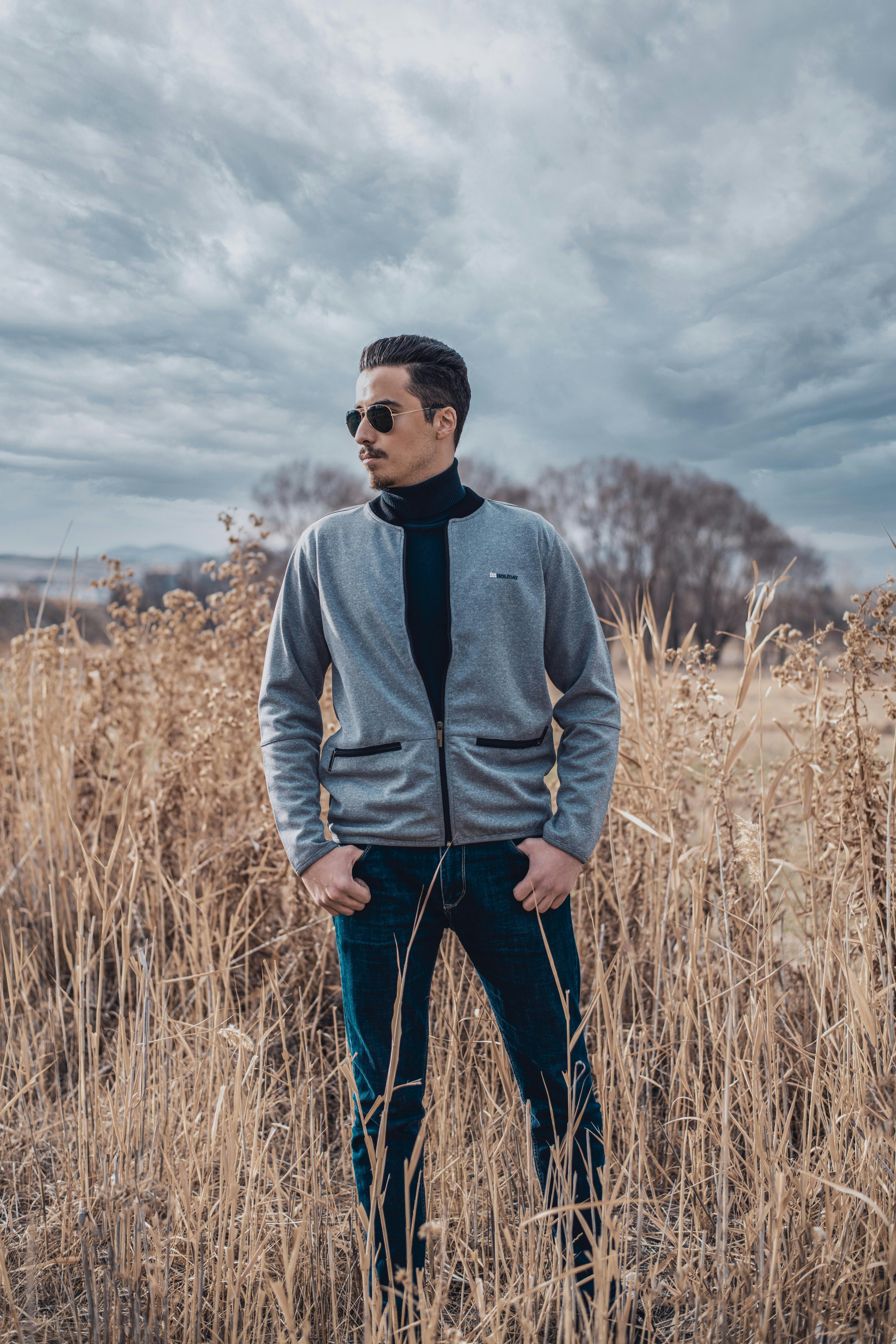 Man in sunglasses and a stylish gray jacket stands confidently in a field of tall, golden grass against a moody sky.