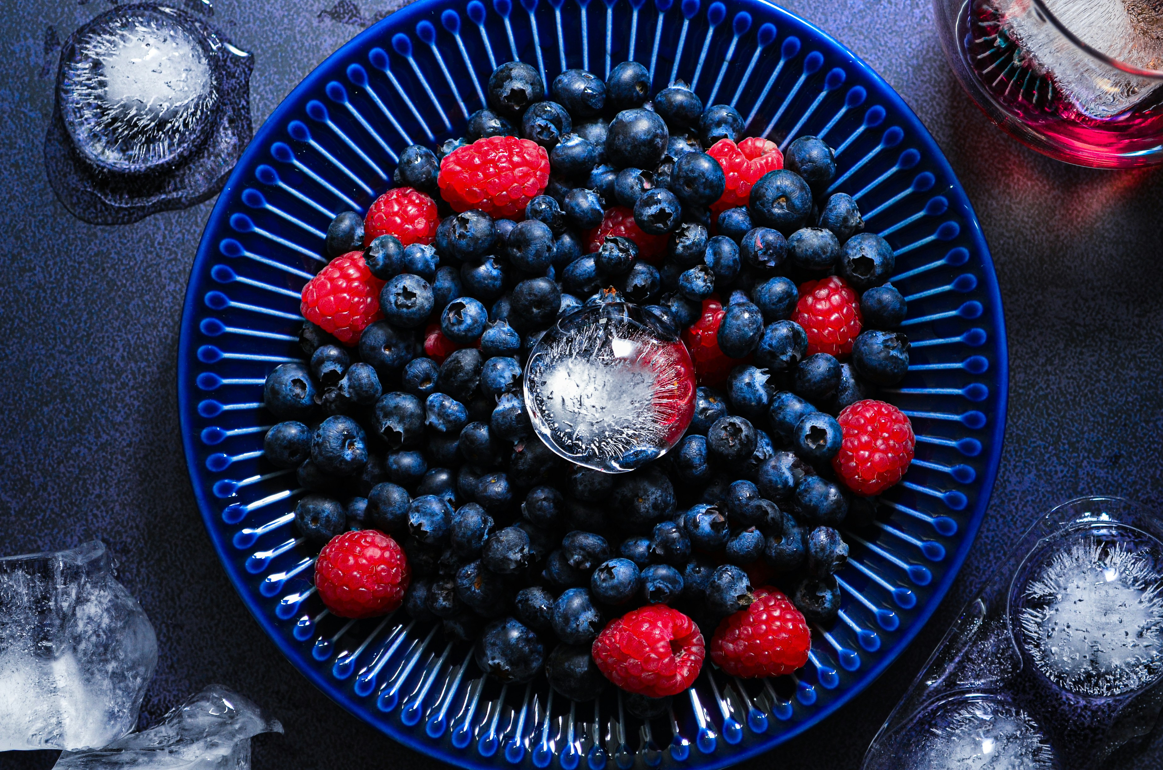A vibrant arrangement of blueberries and raspberries artfully displayed in a blue bowl, surrounded by scattered ice cubes. The composition highlights freshness and summer vibes.