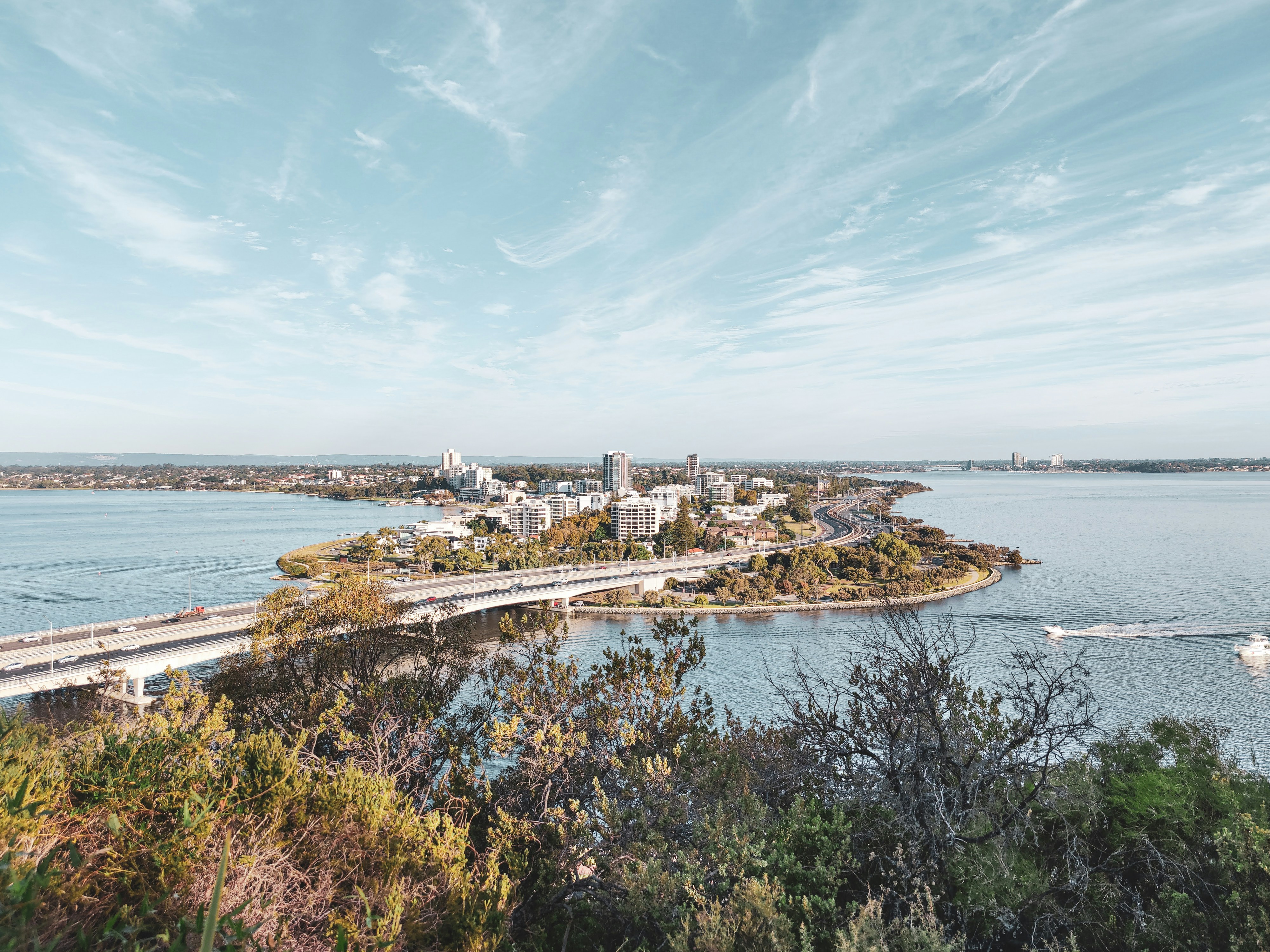 Cityscape with a bridge spanning a river, framed by lush foliage and expansive sky.