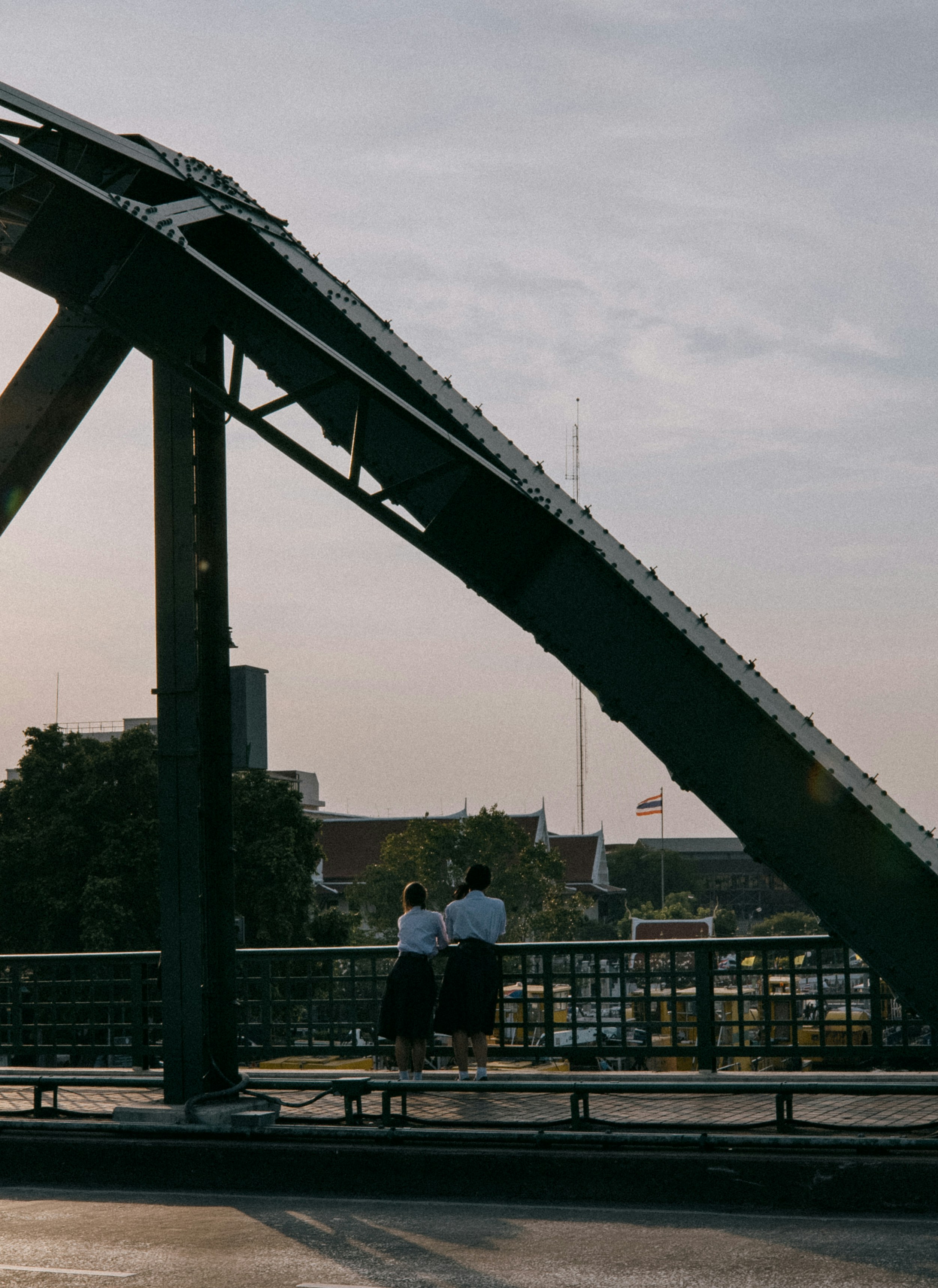 Two people stand at a railing beneath a diagonal steel bridge. The city skyline and a muted sunset provide a calm urban backdrop.