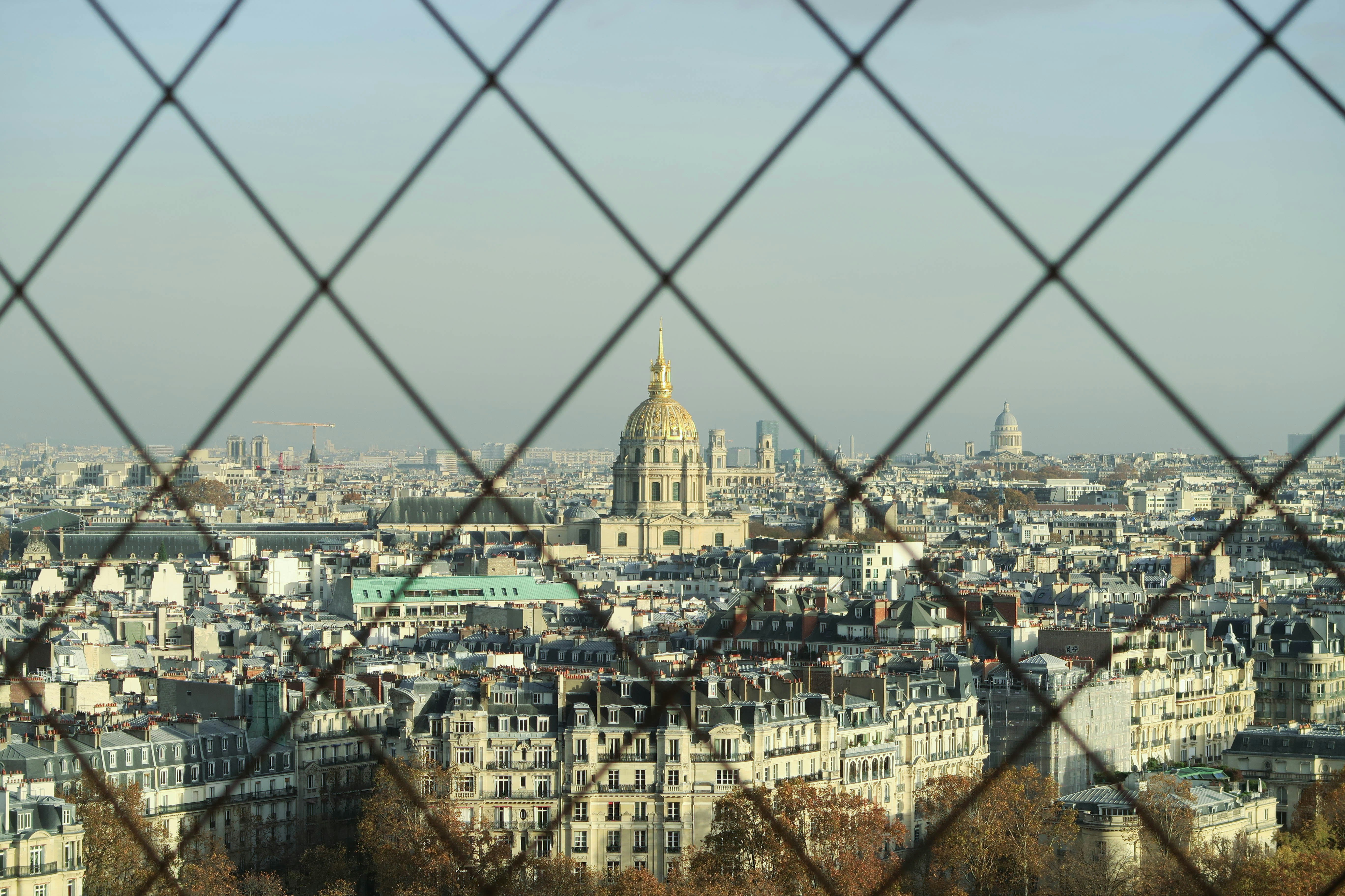 Golden dome of Les Invalides viewed through a diamond-patterned fence, showcasing the sprawling Parisian rooftops below.