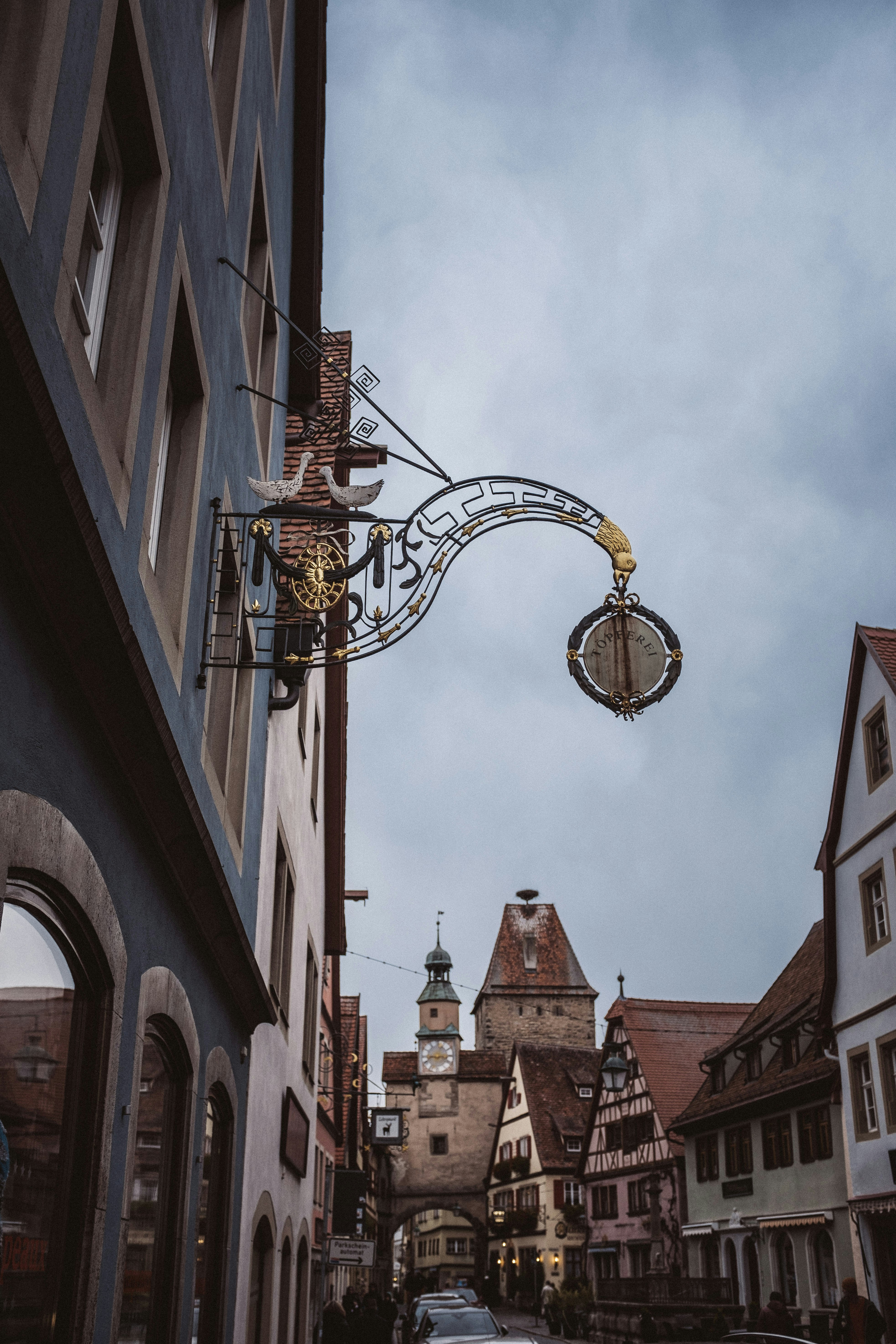 Intricate wrought-iron sign hangs above a cobblestone street, leading to a historic clock tower amidst charming half-timbered houses.