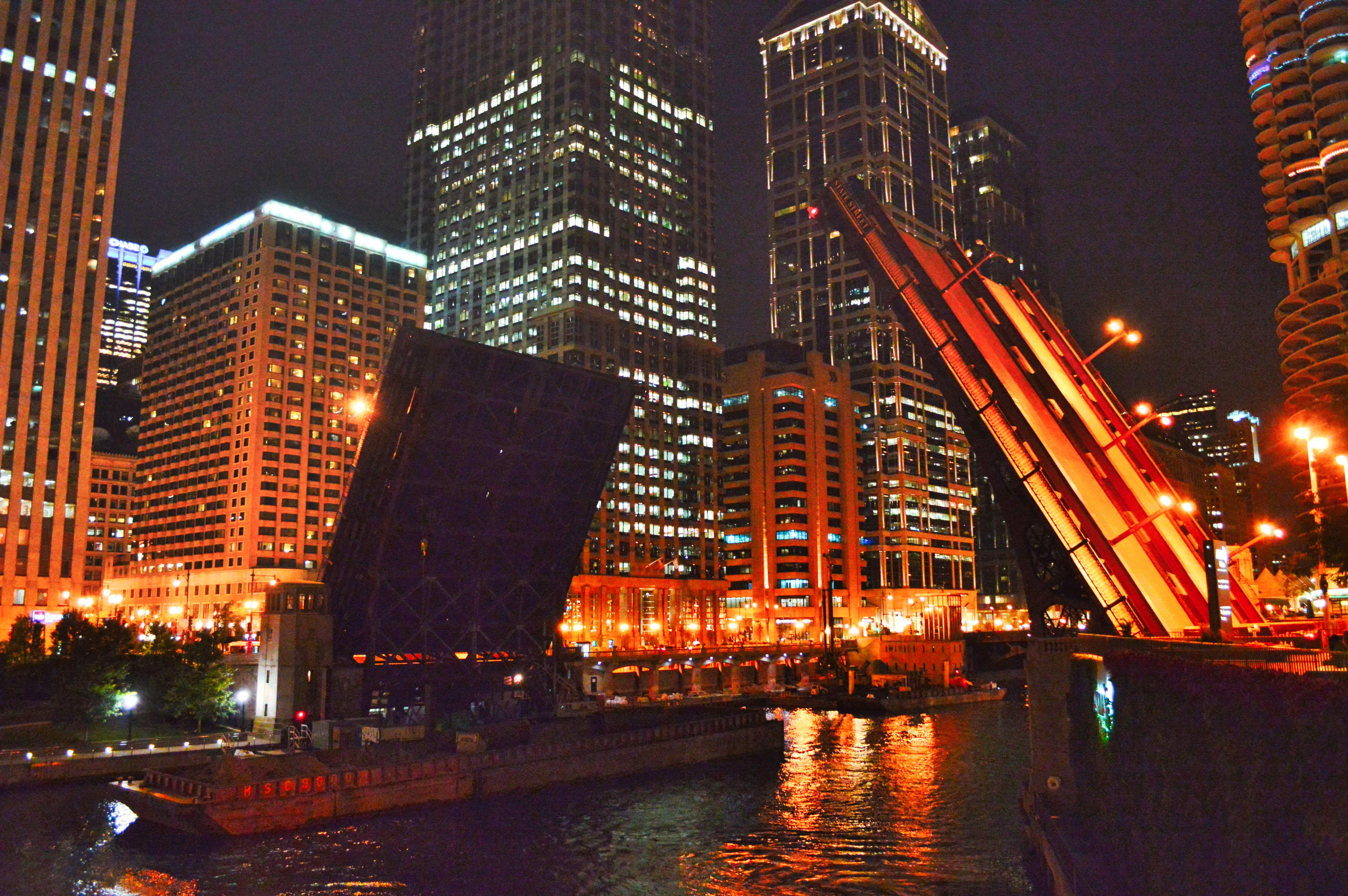 Drawbridge rising against a backdrop of illuminated skyscrapers, reflecting in the water below.