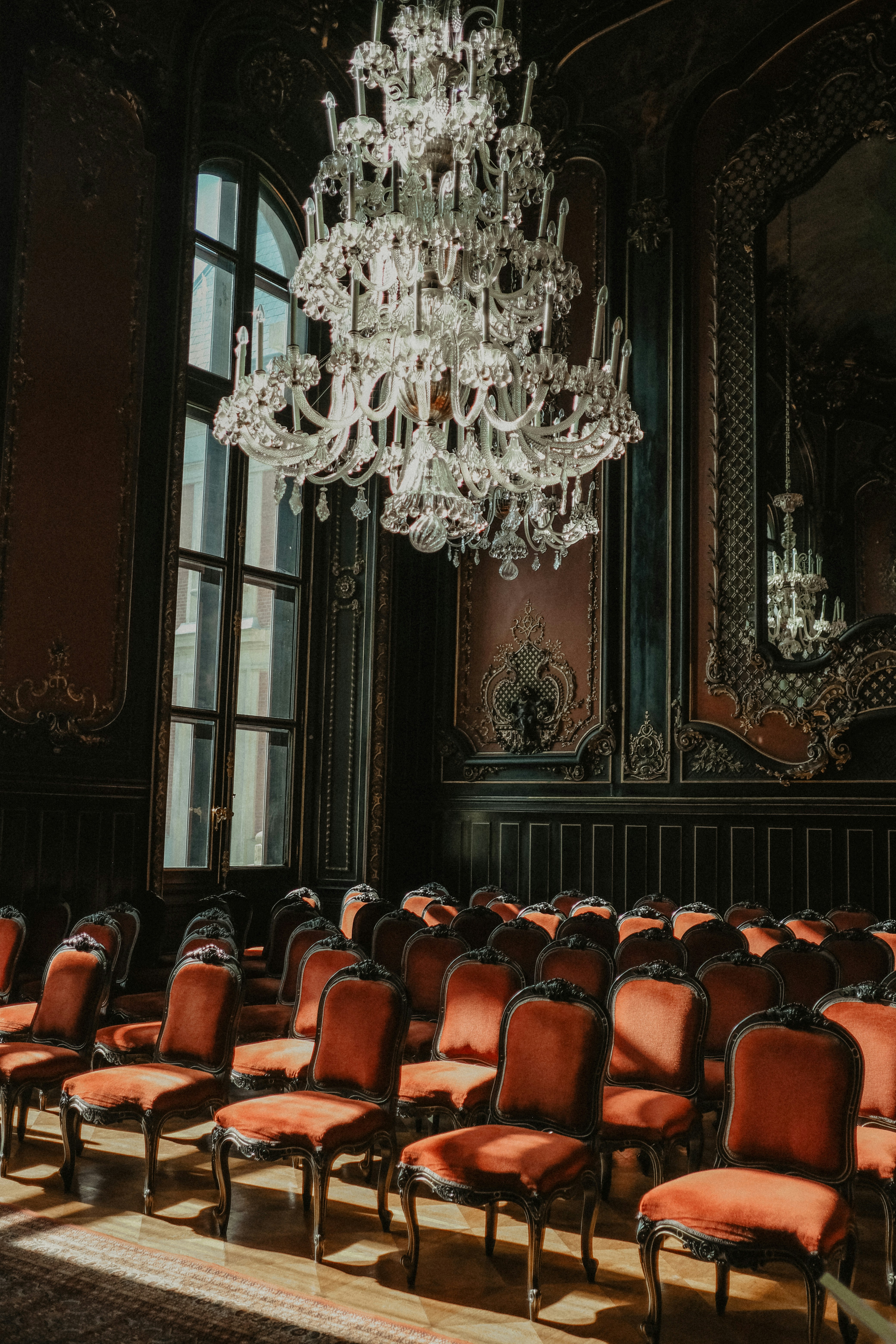 Opulent chandelier hangs above rows of ornate chairs in a lavishly decorated room, showcasing intricate architectural details. 