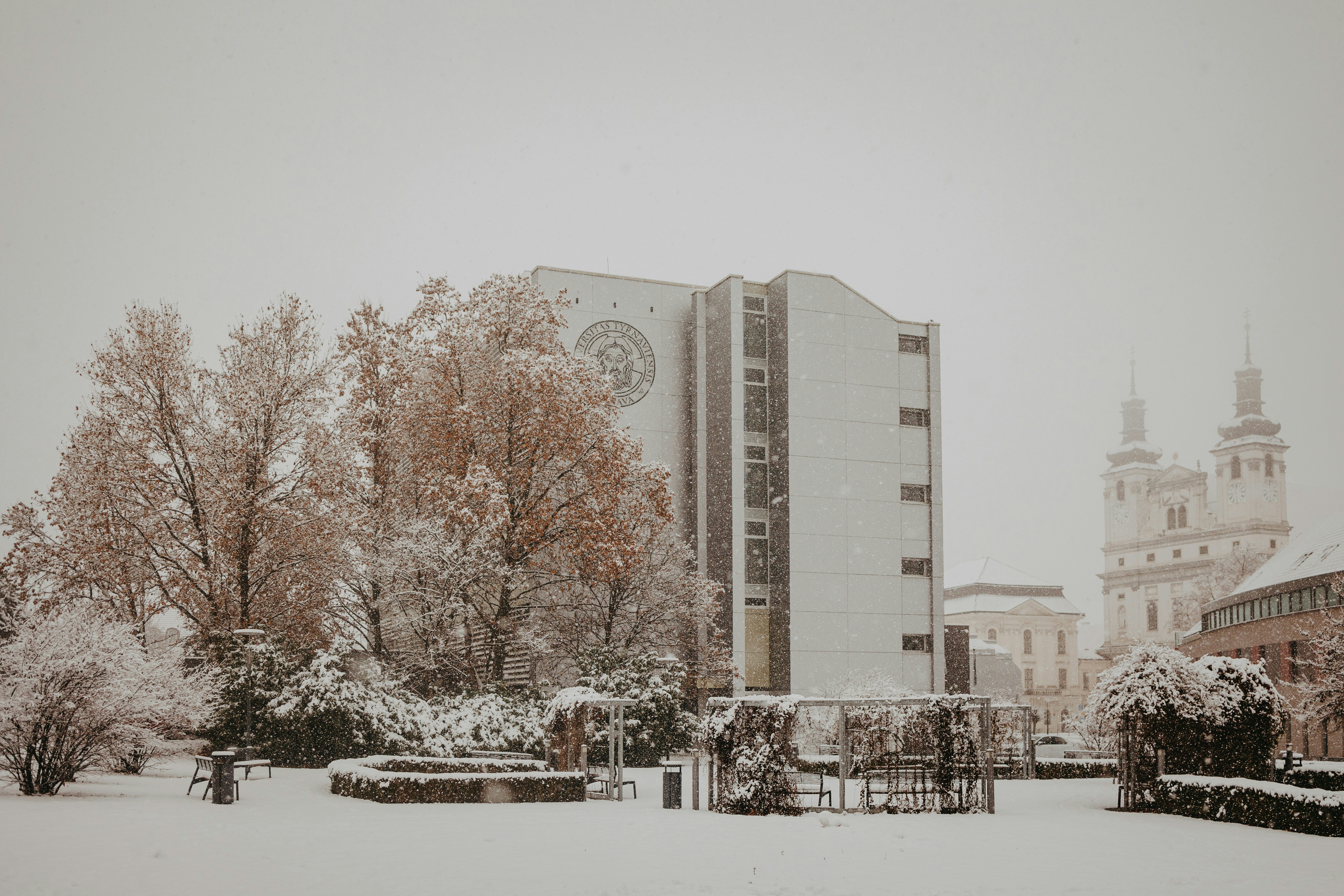 Snow-covered landscape featuring a modern building surrounded by trees with autumn leaves and historical architecture in the background.