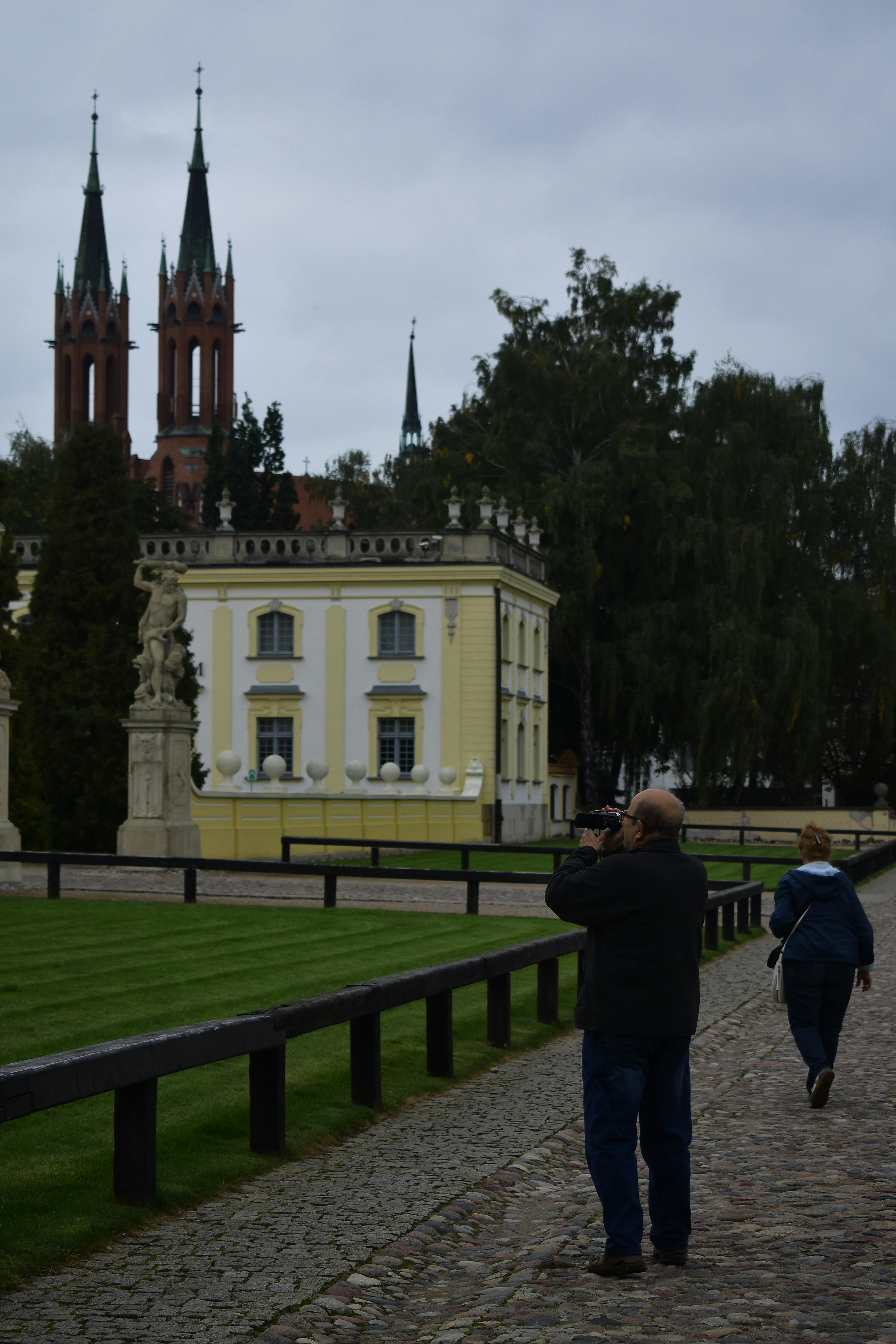Two people walking along a cobblestone path with a view of Gothic church spires and a historic building under a cloudy sky.