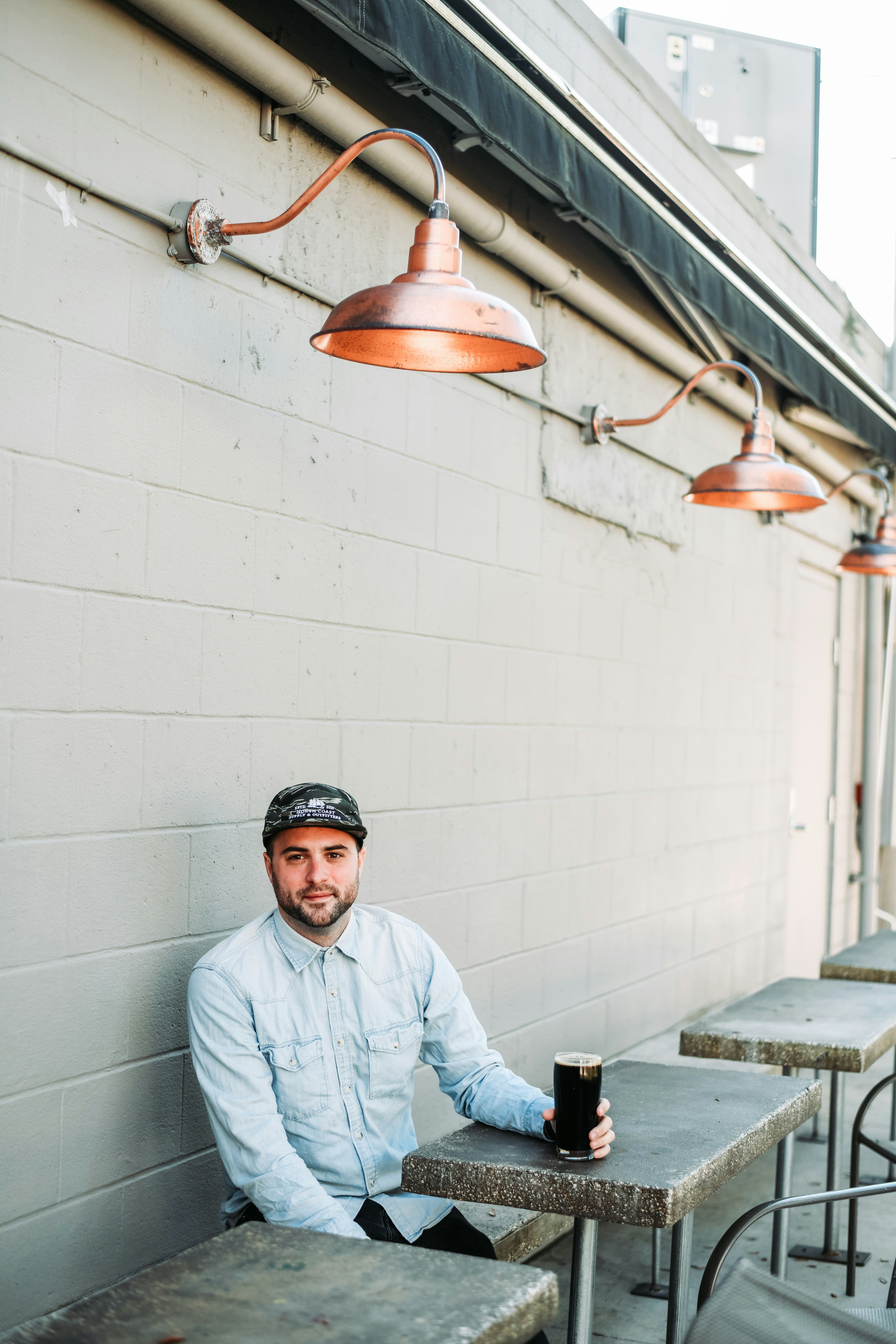 Man sitting at a table with a dark beverage, relaxed against a textured wall with industrial lighting fixtures above.