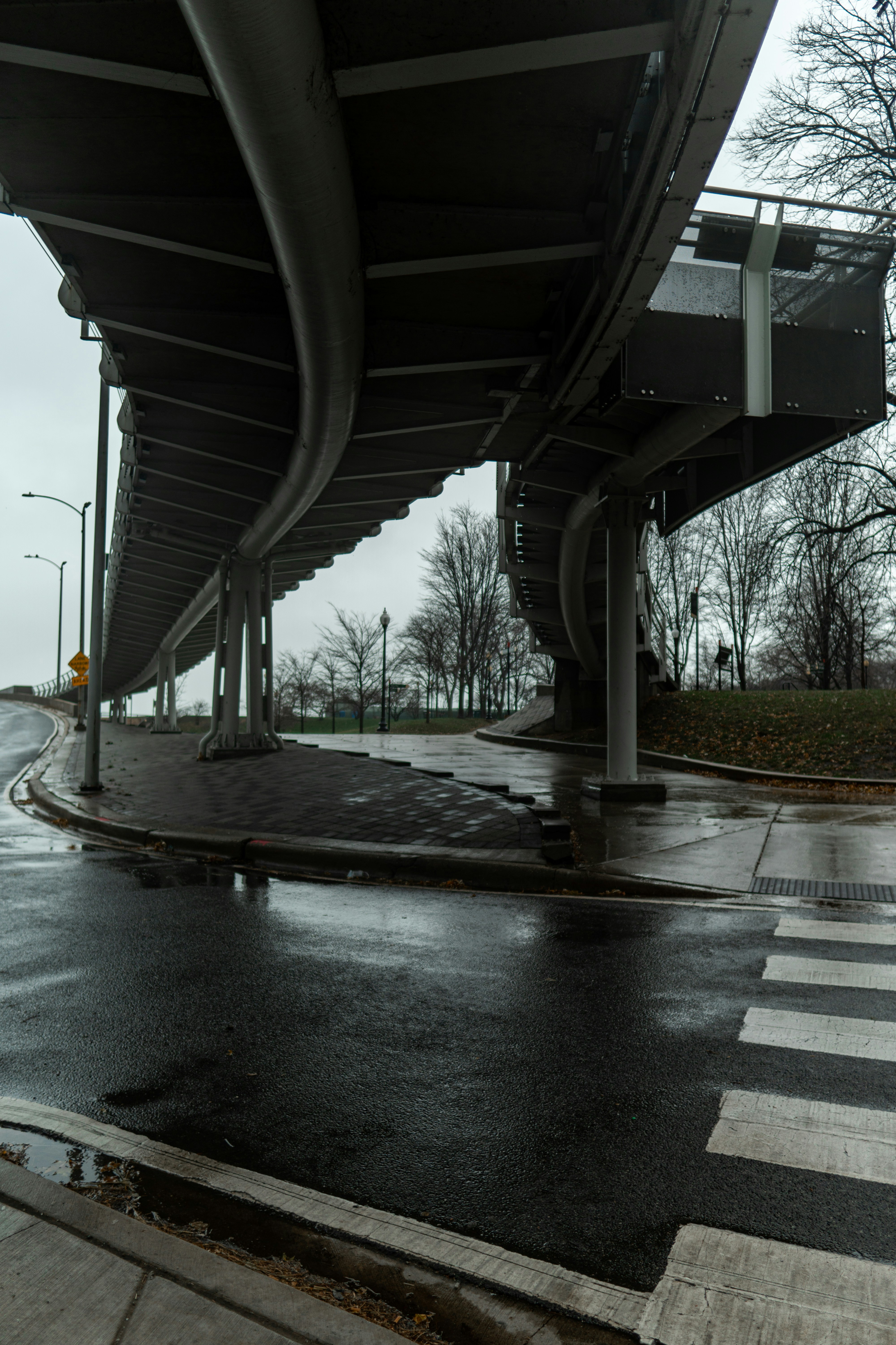Curved overpass structure casting reflections on wet pavement, surrounded by barren trees and a cloudy sky.