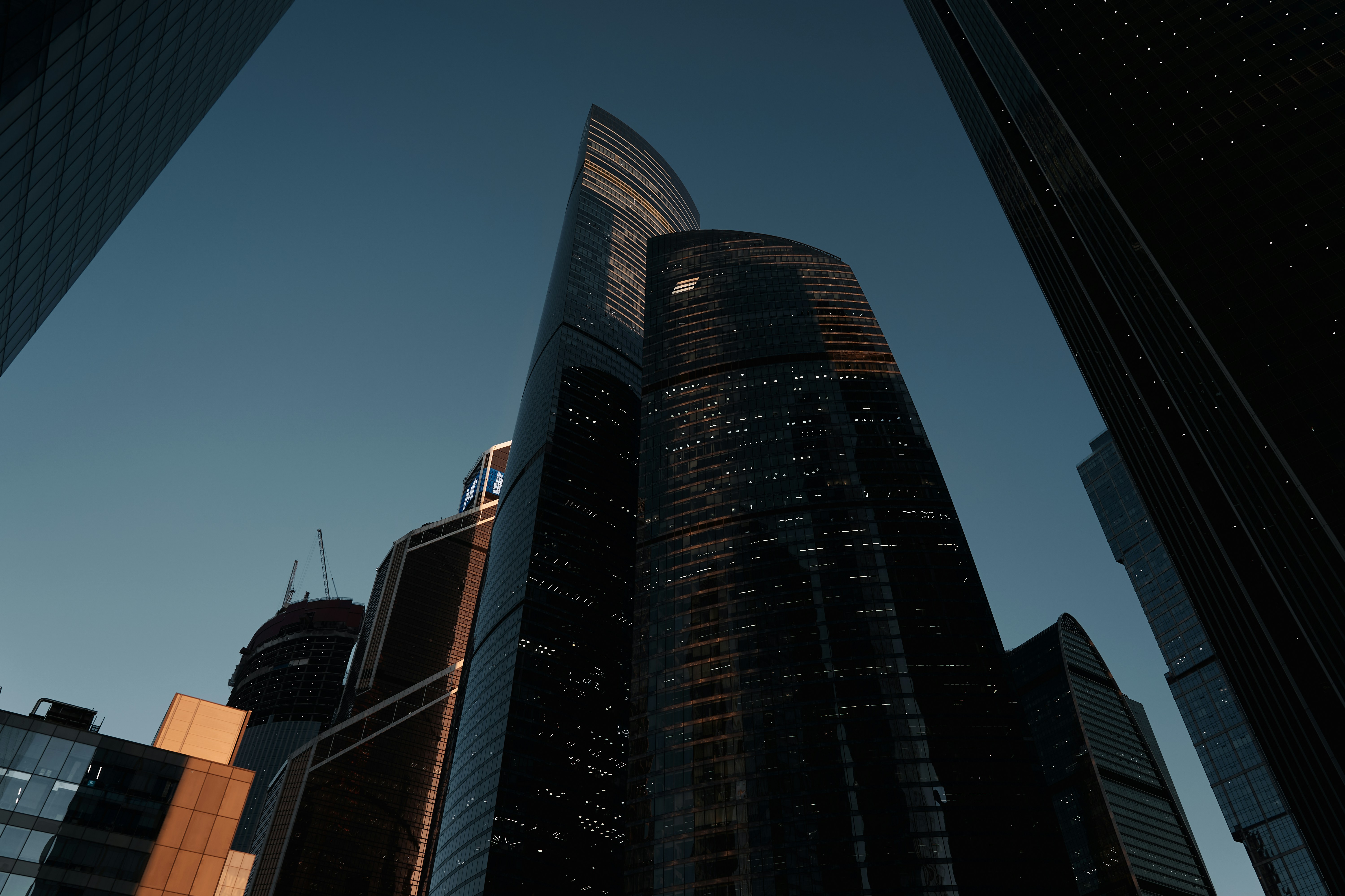 Modern skyscrapers rise against a twilight sky, showcasing reflections of the setting sun and city lights. The architectural forms create a dynamic urban landscape.
