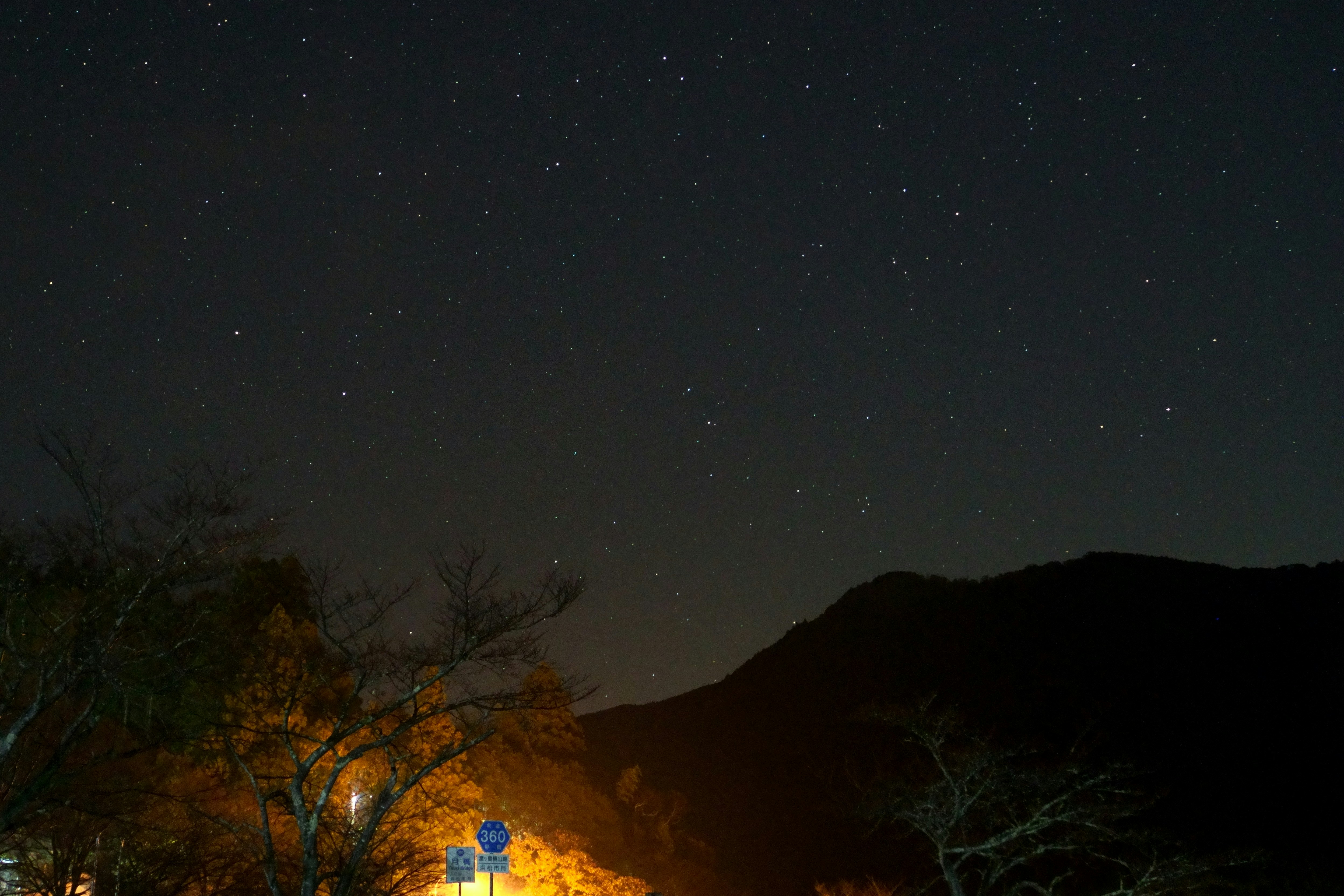 Starry sky illuminating a tranquil landscape, with silhouetted trees and distant mountains. The glow of artificial light adds depth to the serene night scene.