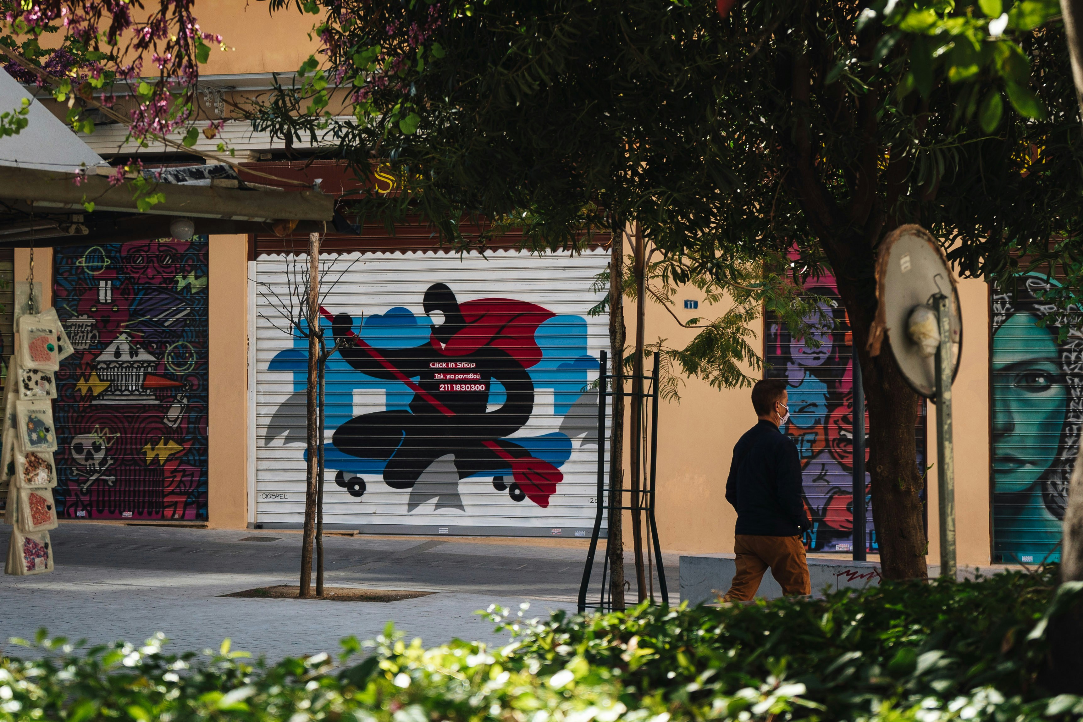 This engaging urban scene captures vibrant street art on a row of shop shutters, featuring bold graffiti with striking hues of blue, red, and black. The composition is accentuated by natural elements like trees and foliage, which frame the image and add depth. The contrast of the colorful artwork against the muted tones of the surrounding environment creates a dynamic and visually striking atmosphere, further enhanced by the play of light and shadow.