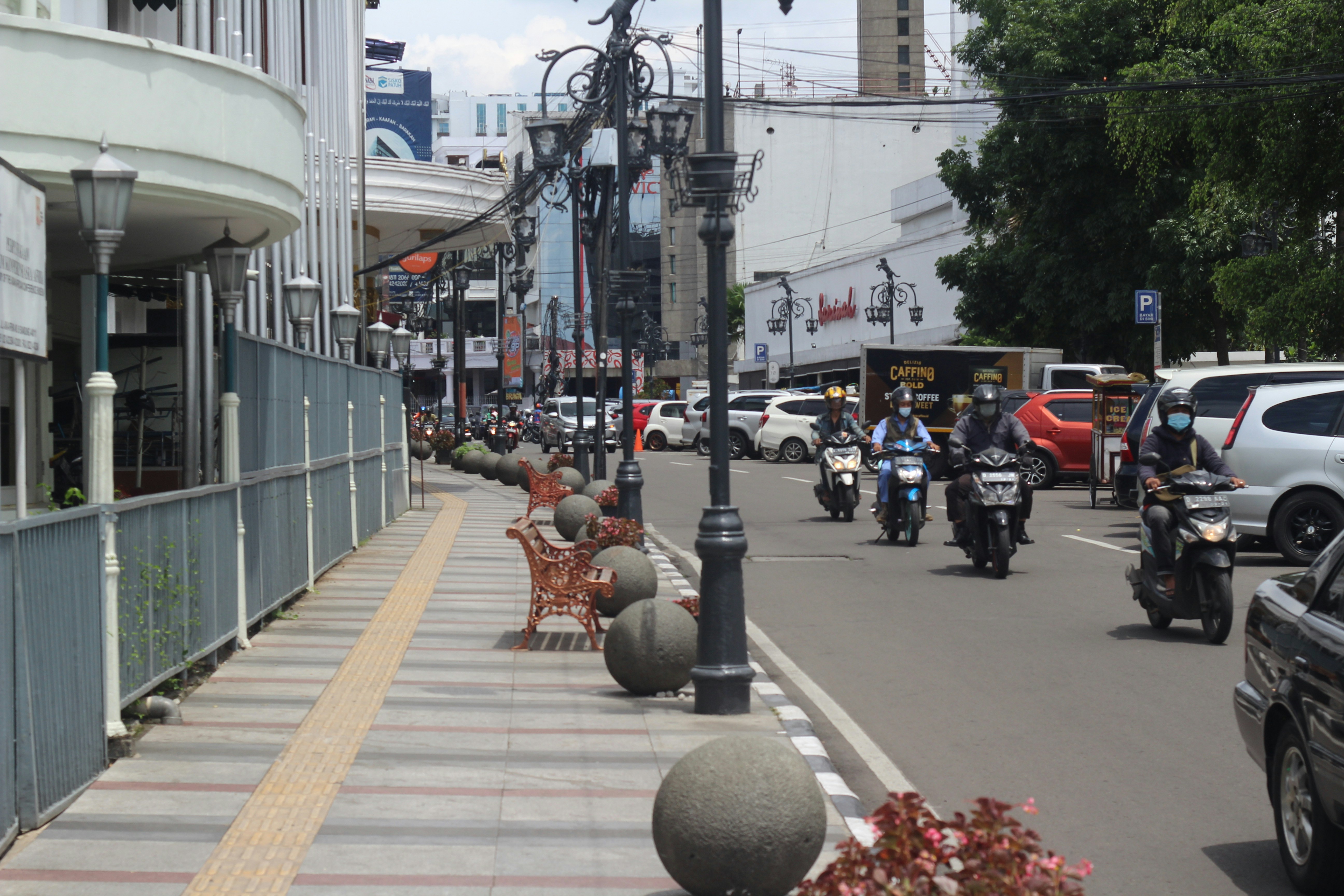 Busy city street with motorcycles and cars alongside a pedestrian walkway lined with spherical bollards.