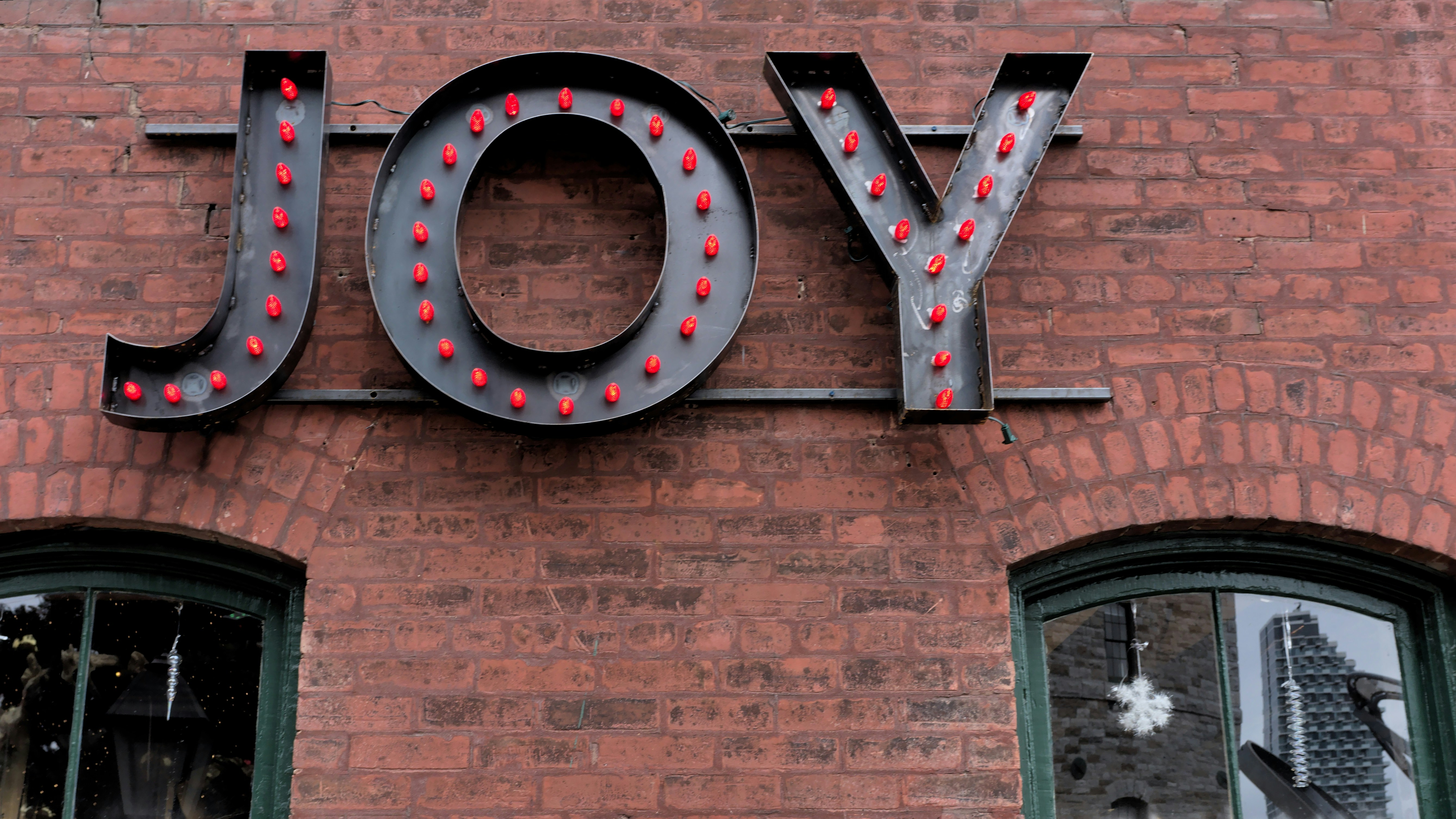 Metallic letters spelling 'JOY' adorned with bright red bulbs against a rustic brick wall.