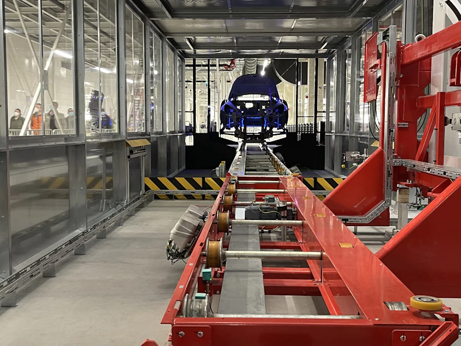 Automated assembly line in a metalwork facility with a blue vehicle resting on the distant track, framed by red support structures and glass-walled observation bays.