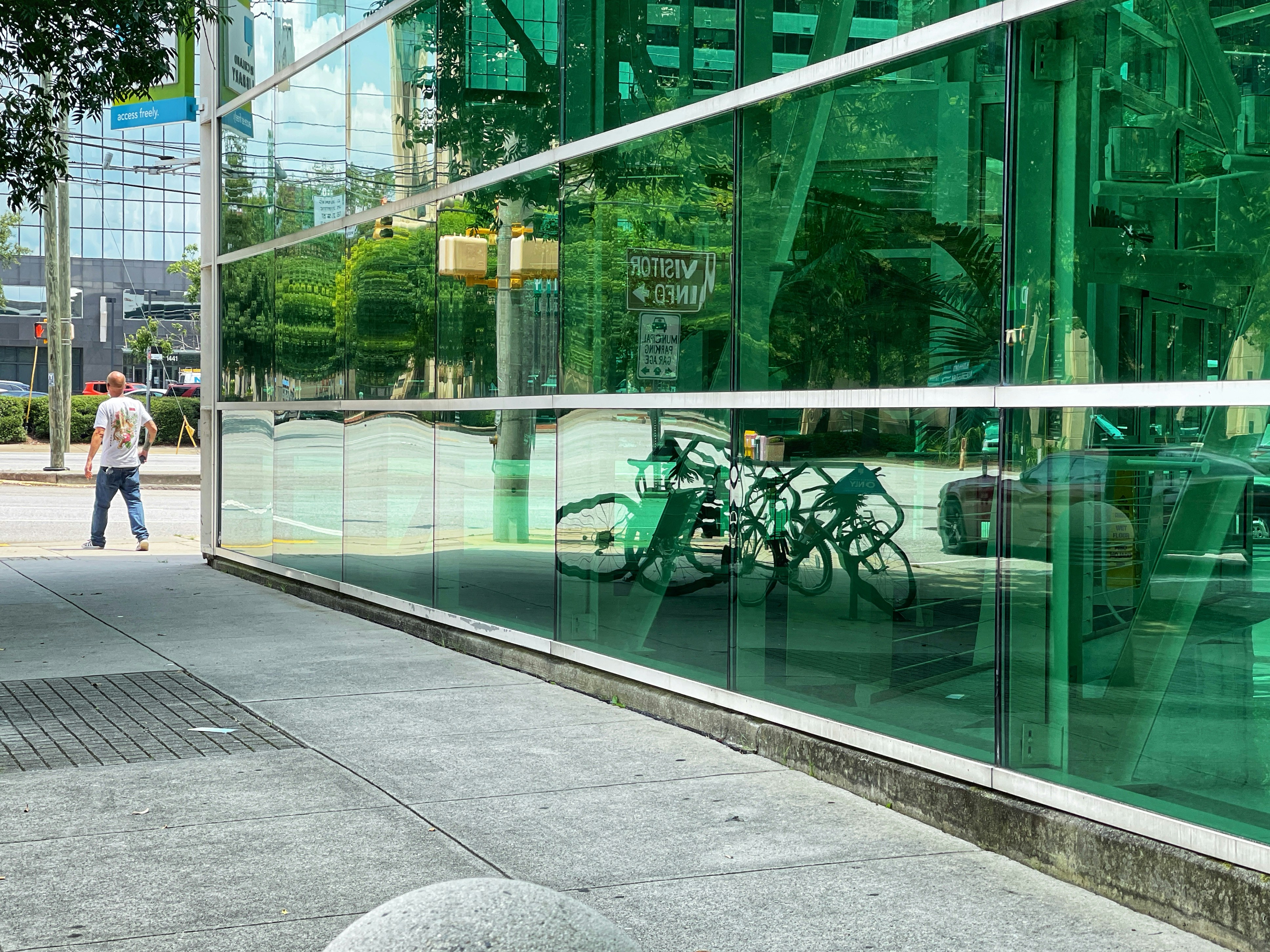 Reflections of bicycles and surrounding greenery on the green-tinted glass facade of a modern building.