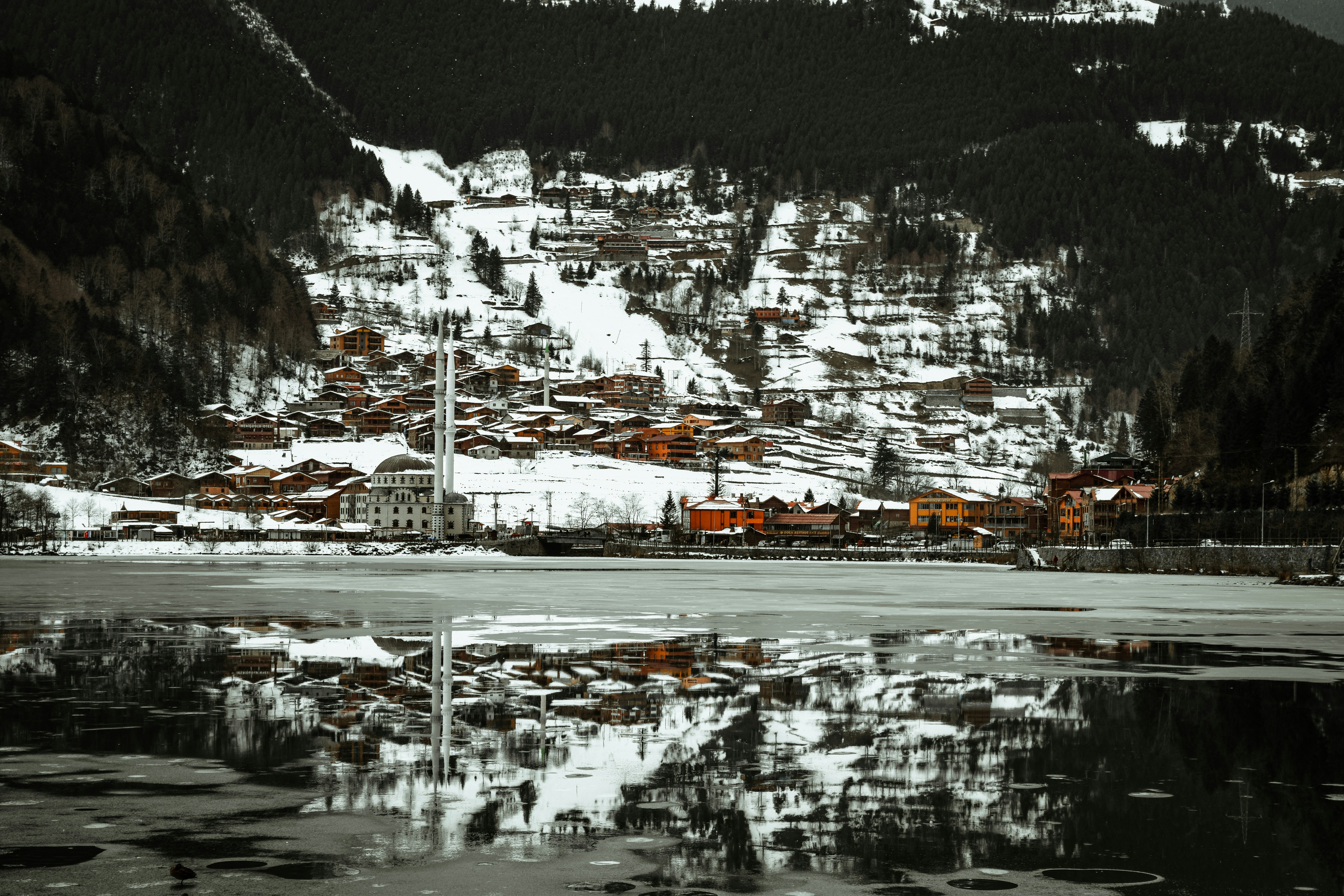 Snow-covered mountain village reflected on a tranquil icy lake under a cloudy sky.