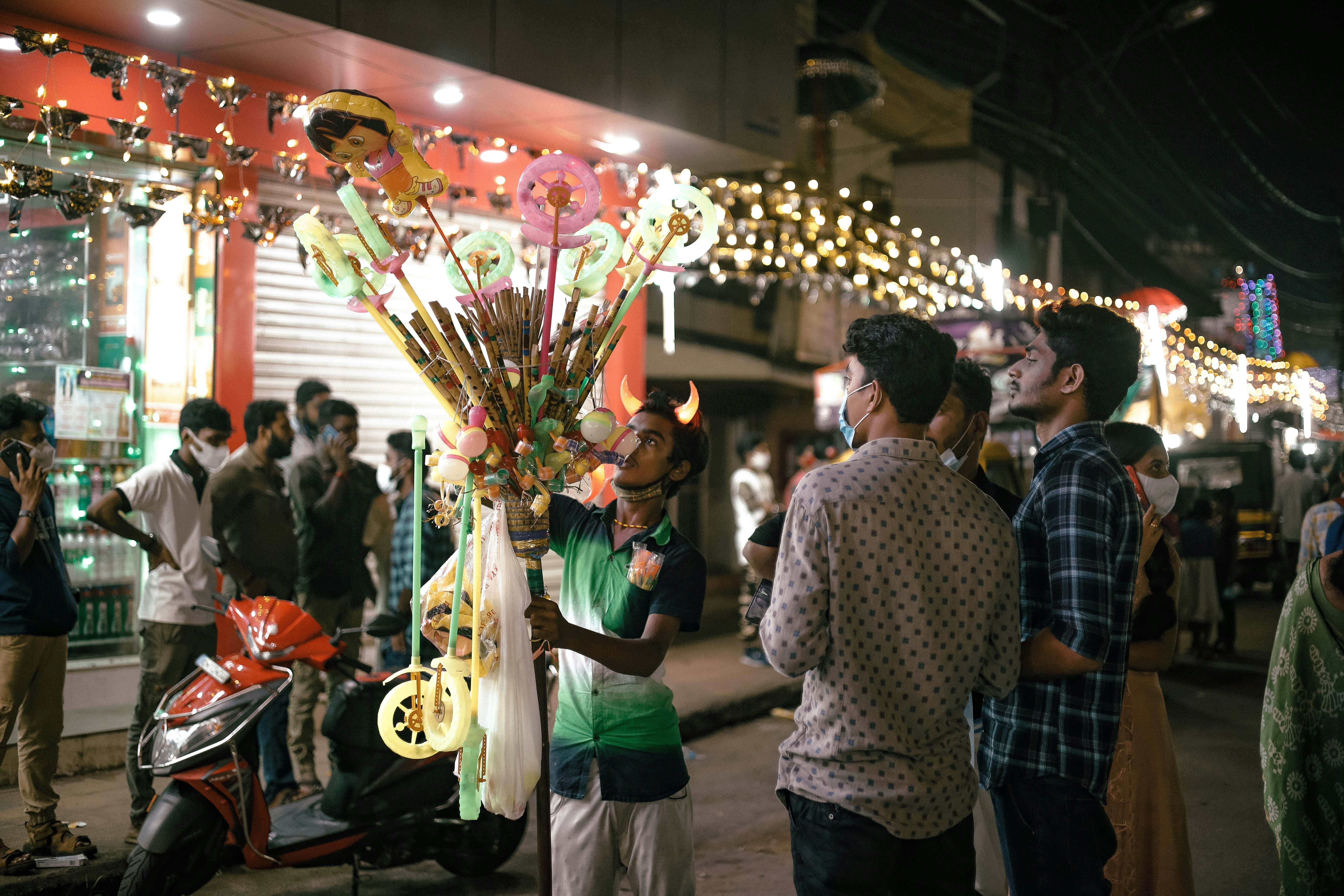 A young vendor showcases a colorful assortment of toys in a bustling night market, surrounded by intrigued onlookers. Bright lights and festive decorations enhance the lively atmosphere.