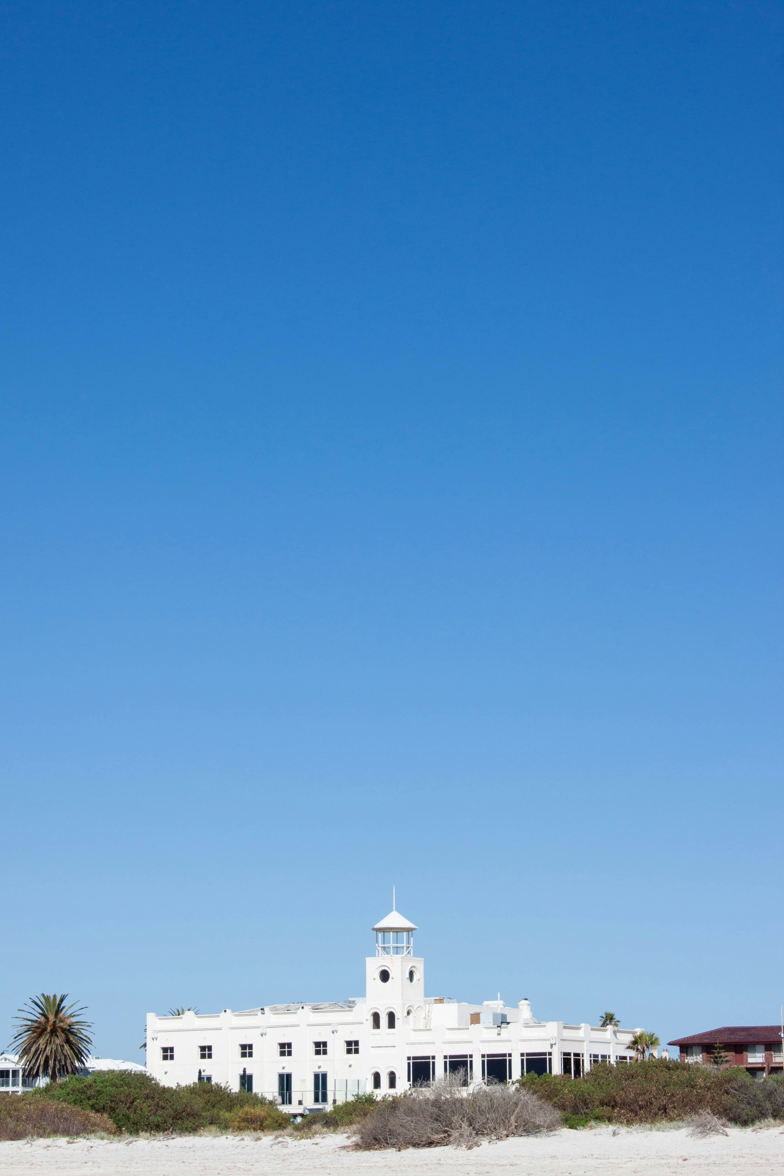 Historic white building stands against a clear blue sky, framed by lush greenery and palm trees.