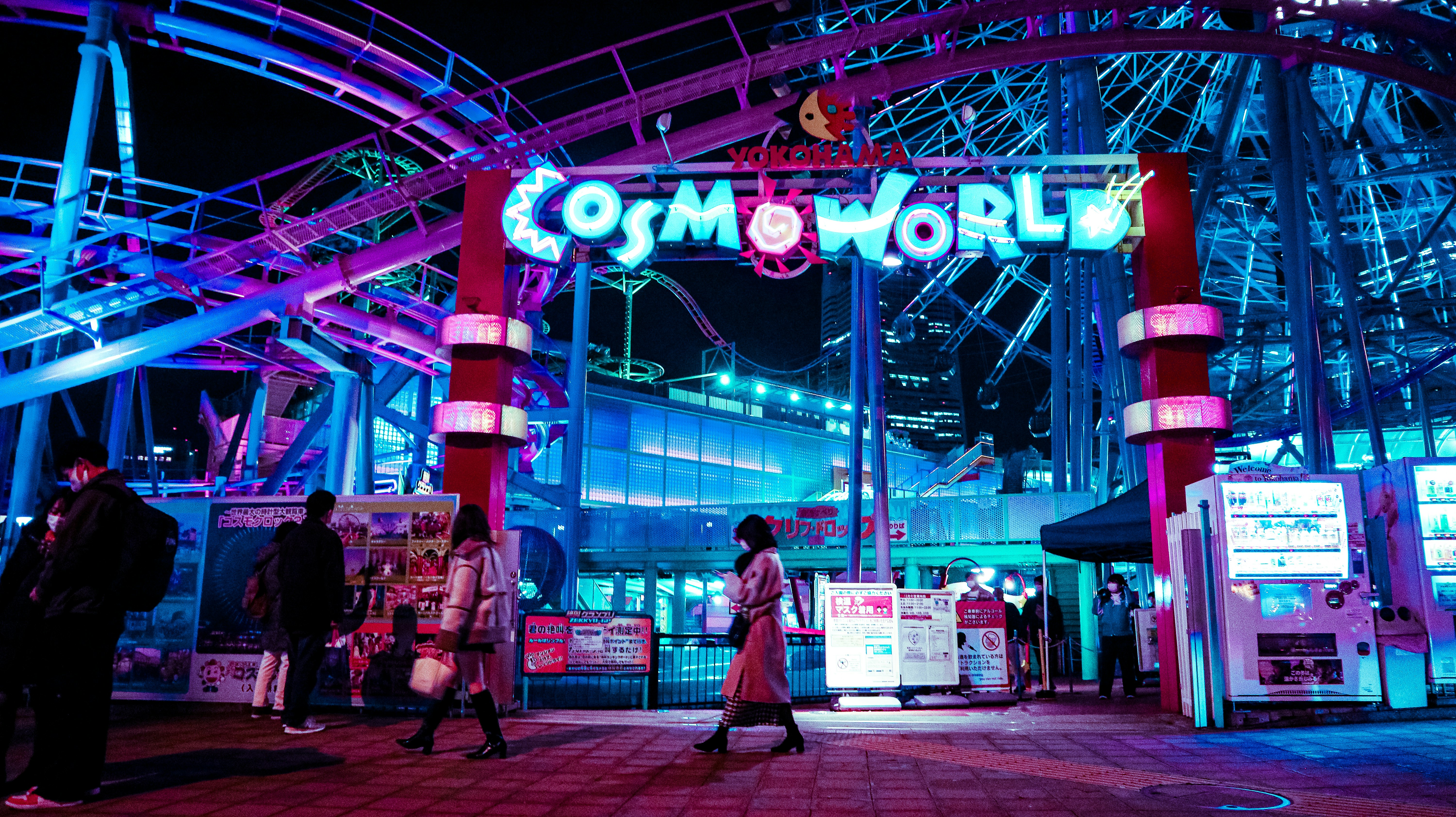 Vibrant neon lights illuminate the entrance of an amusement park with people walking underneath.