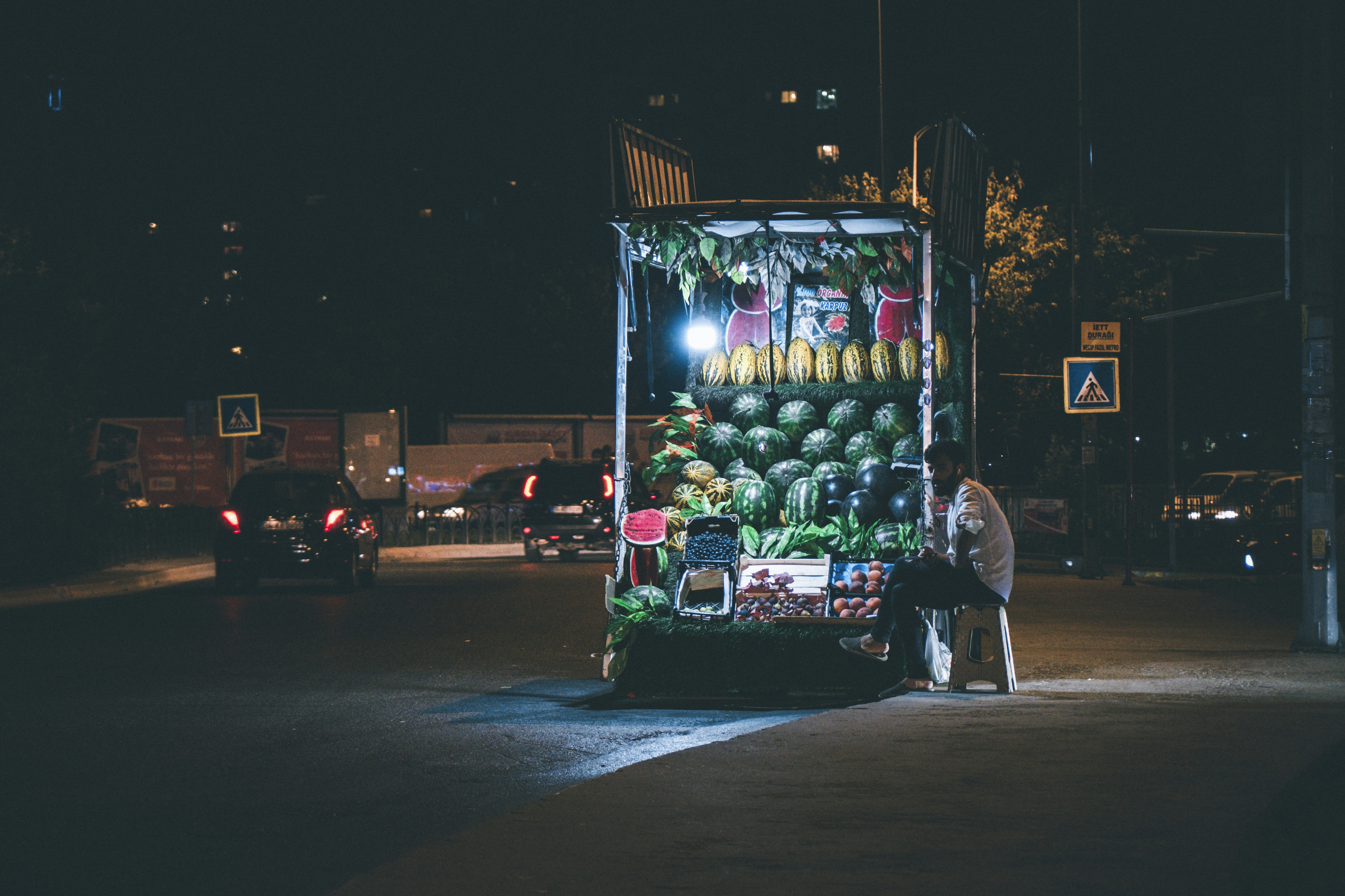 Vibrant fruit stand illuminated at night, showcasing a variety of fresh produce. A vendor sits quietly, surrounded by the colorful display.