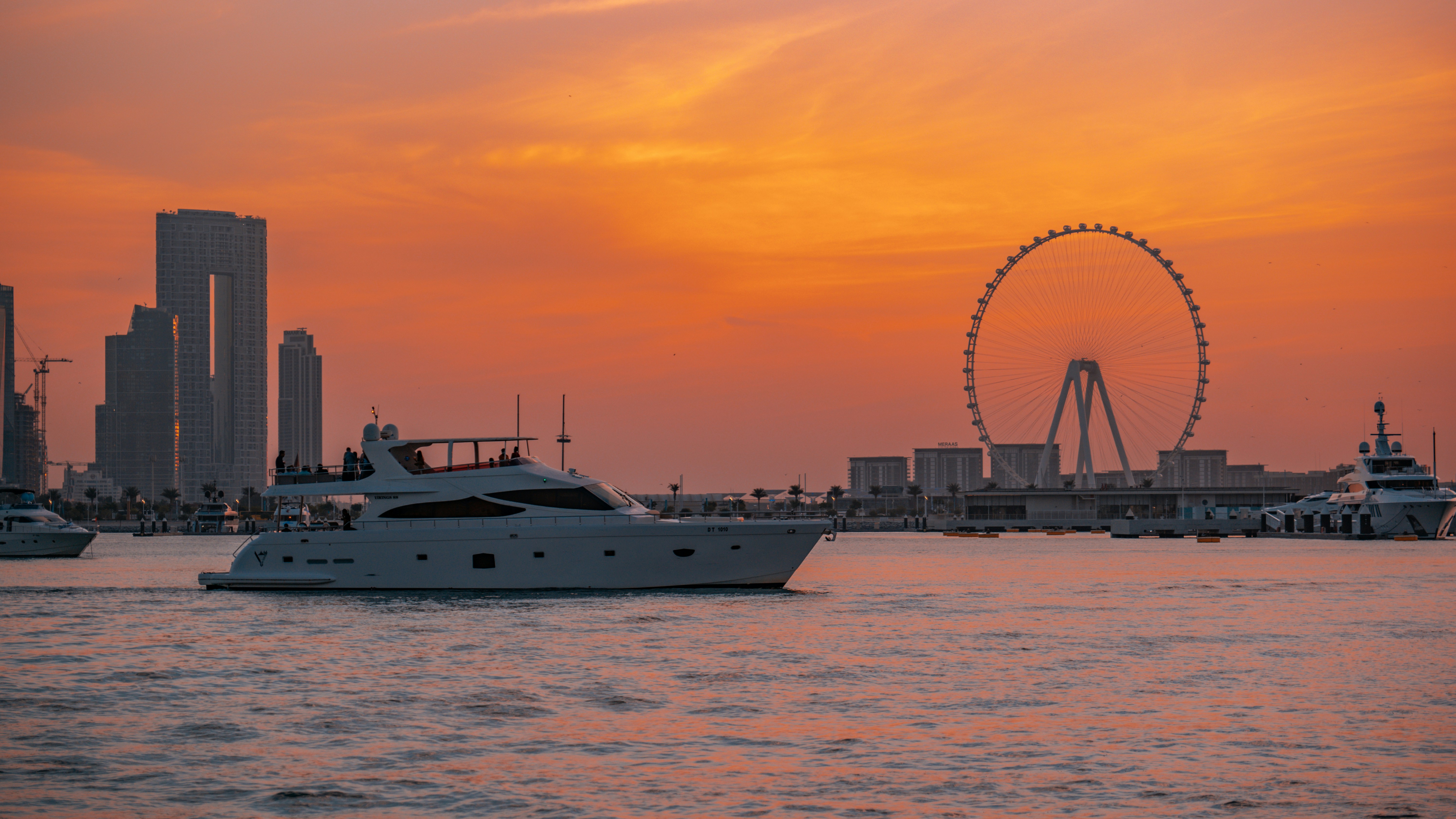 Yacht gliding on a tranquil waterway with a city skyline and Ferris wheel silhouetted against a vibrant orange sunset.