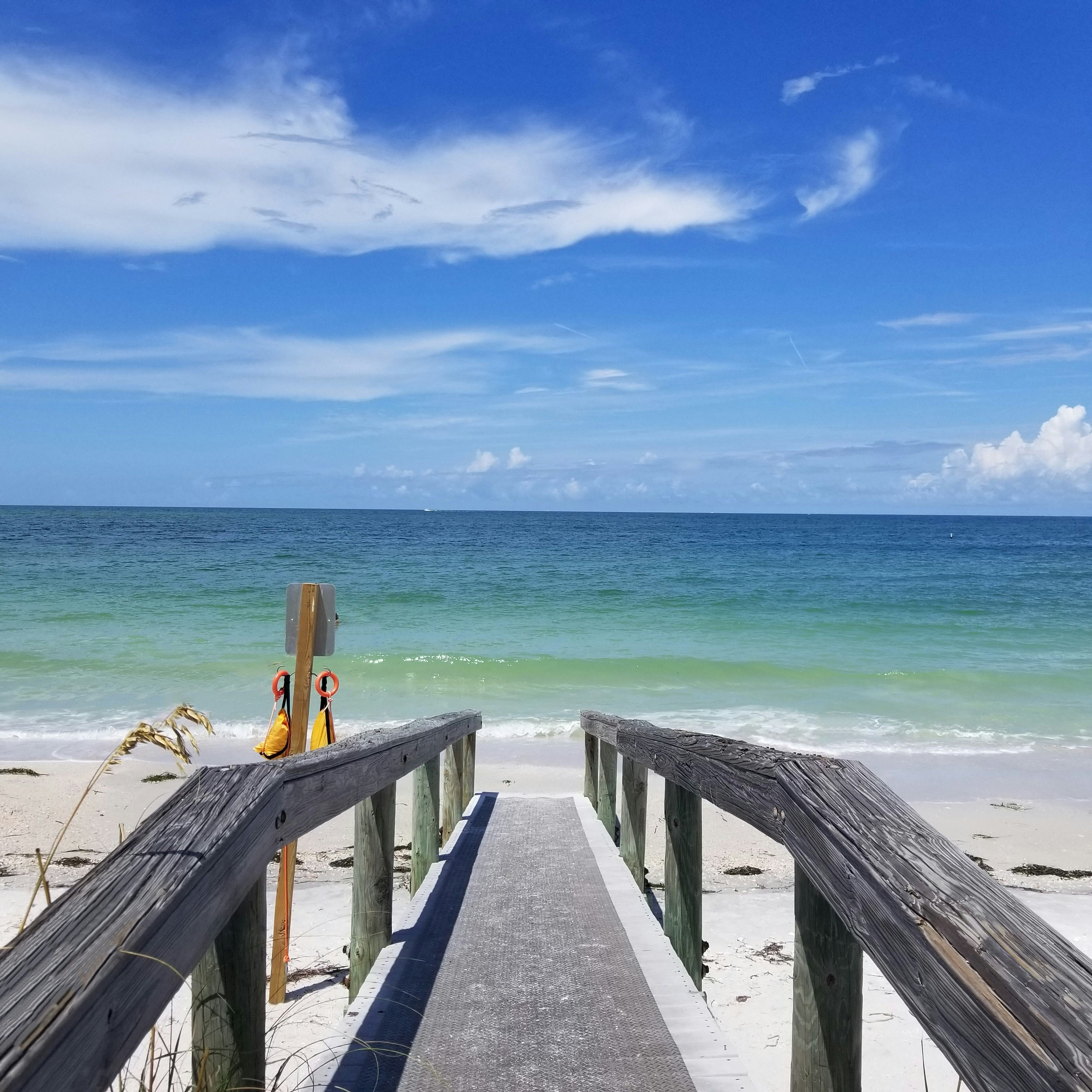 Wooden boardwalk leading to a serene beach with gentle waves and a clear blue sky.