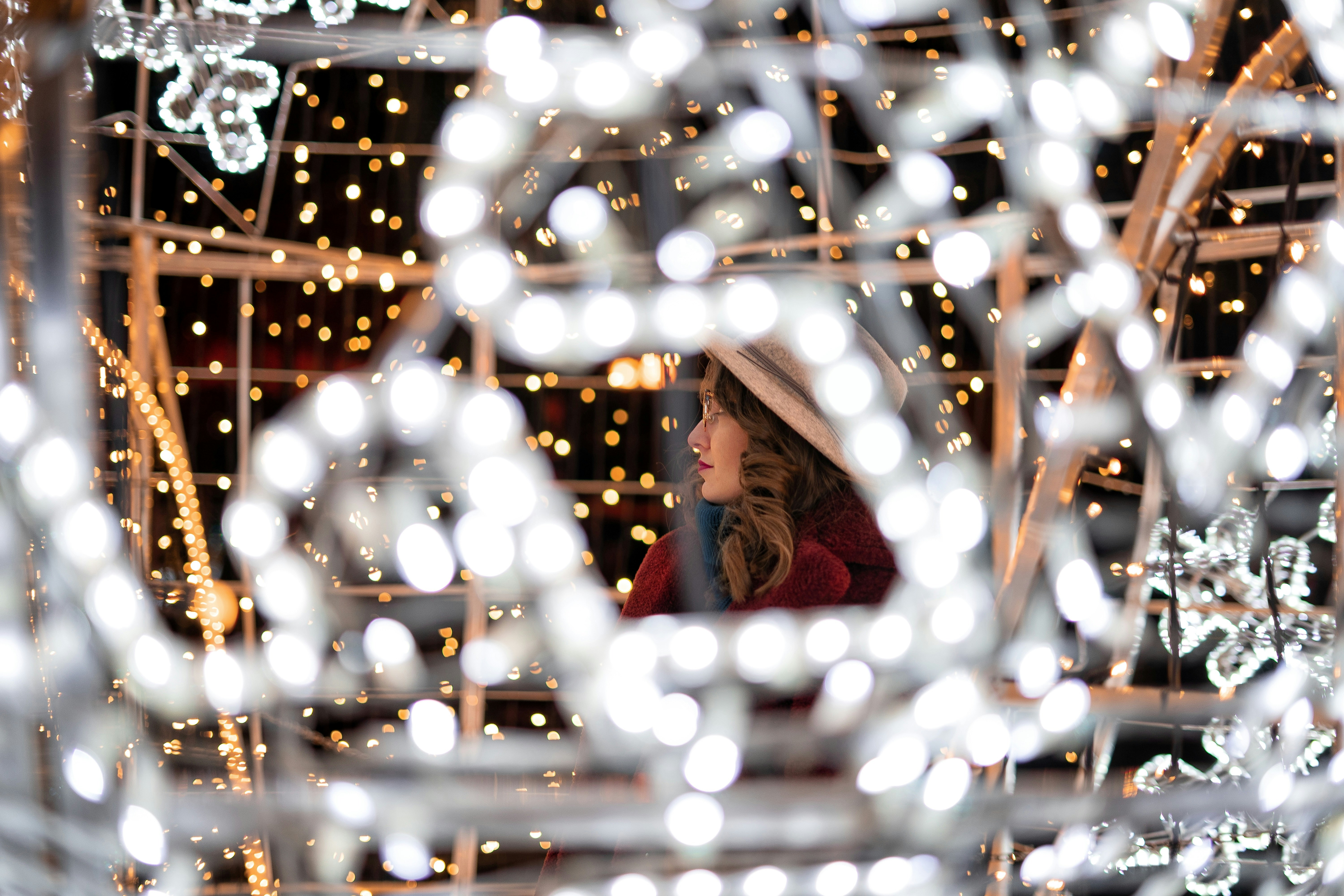 A woman in a cozy hat gazes thoughtfully amidst a backdrop of twinkling holiday lights, framed by a decorative light structure.