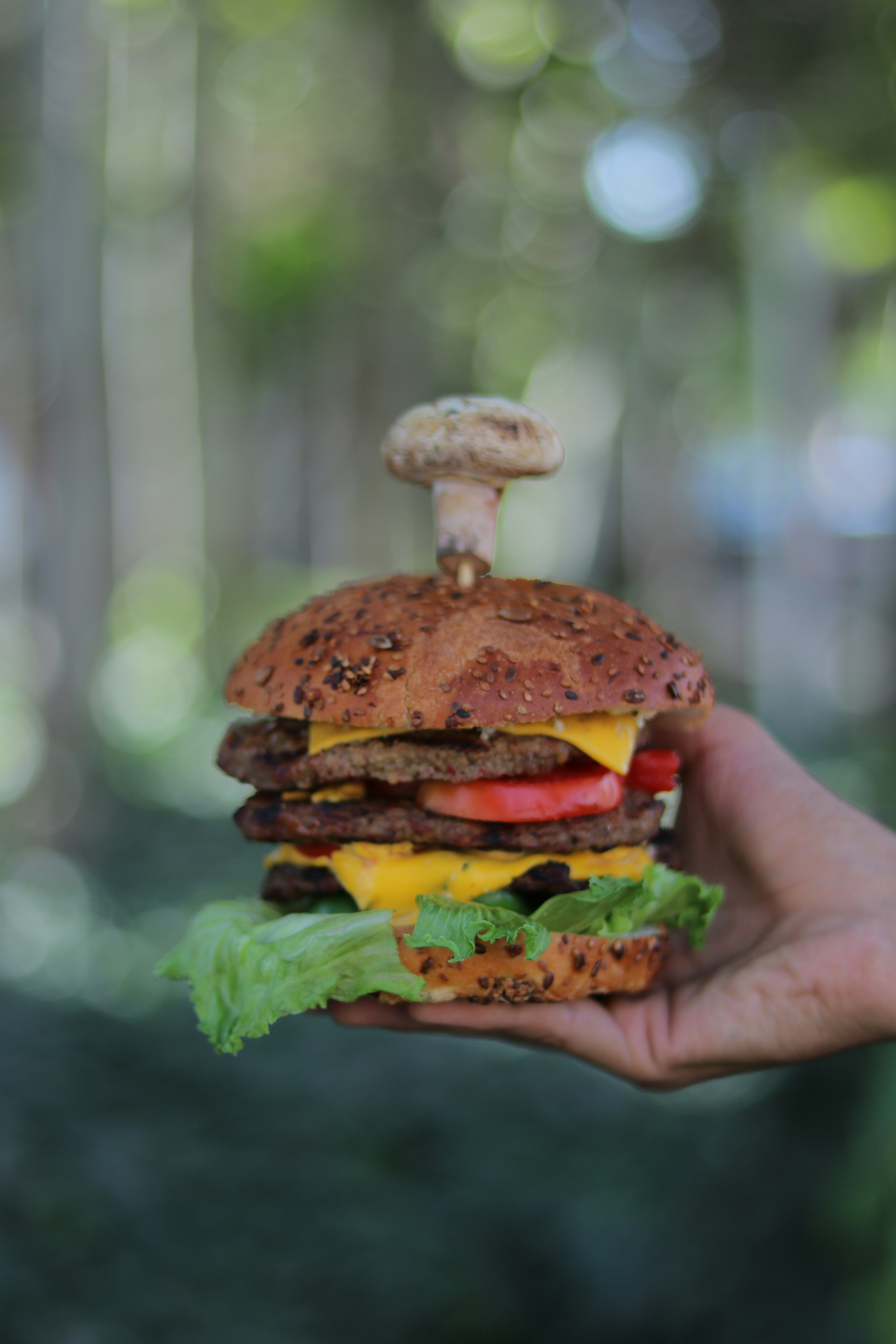 A whimsical burger stacked high with layers of lettuce, tomato, cheese, and patties, topped with a mushroom, held by a hand against a blurred natural backdrop.