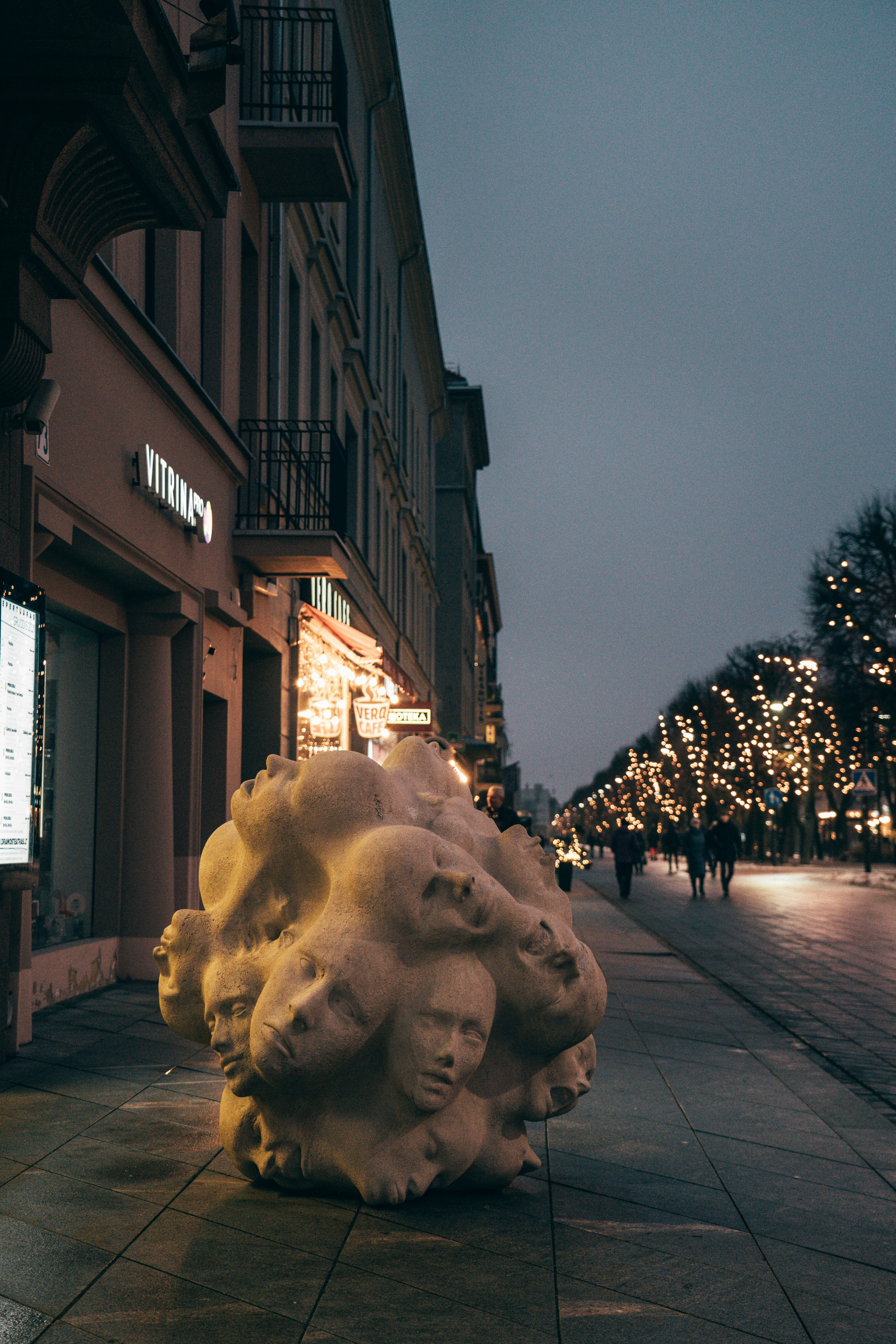 Abstract sculpture featuring multiple human faces, positioned on a city street adorned with festive lights. The scene captures a blend of urban life and artistic expression.