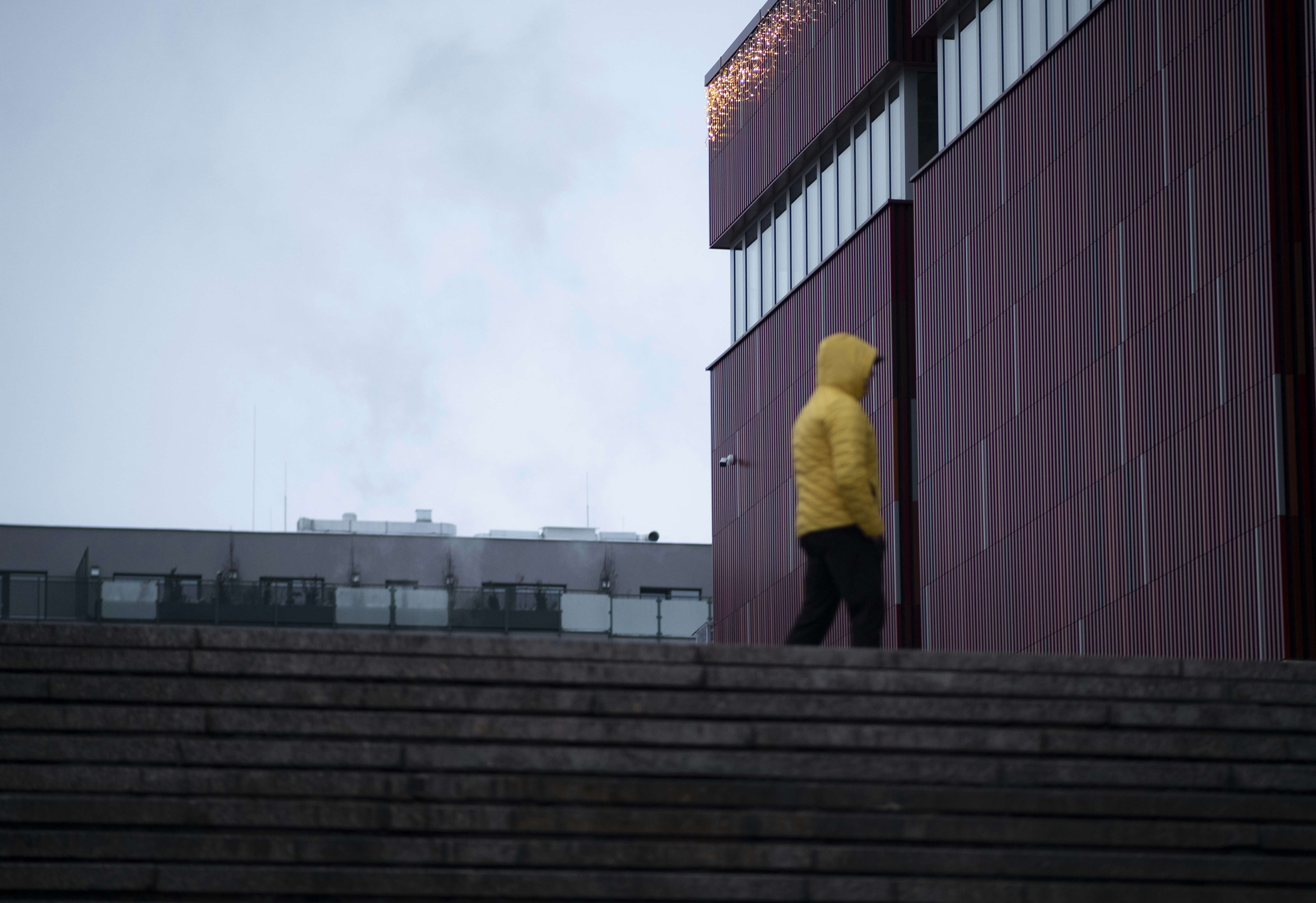 Person in a yellow raincoat walking past a modern building on a cloudy day.