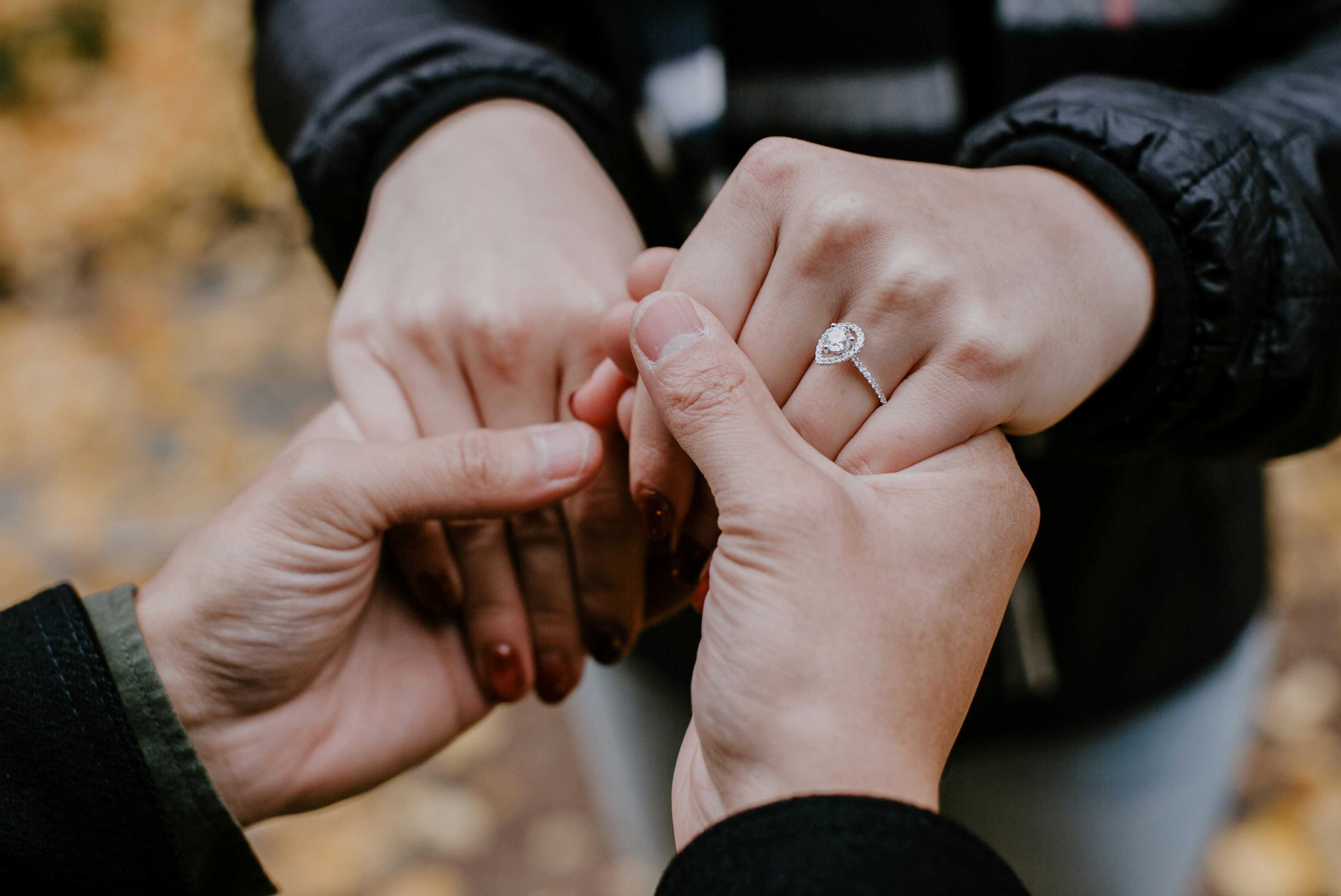 Two hands clasping each other, showcasing an engagement ring amidst a backdrop of autumn leaves.