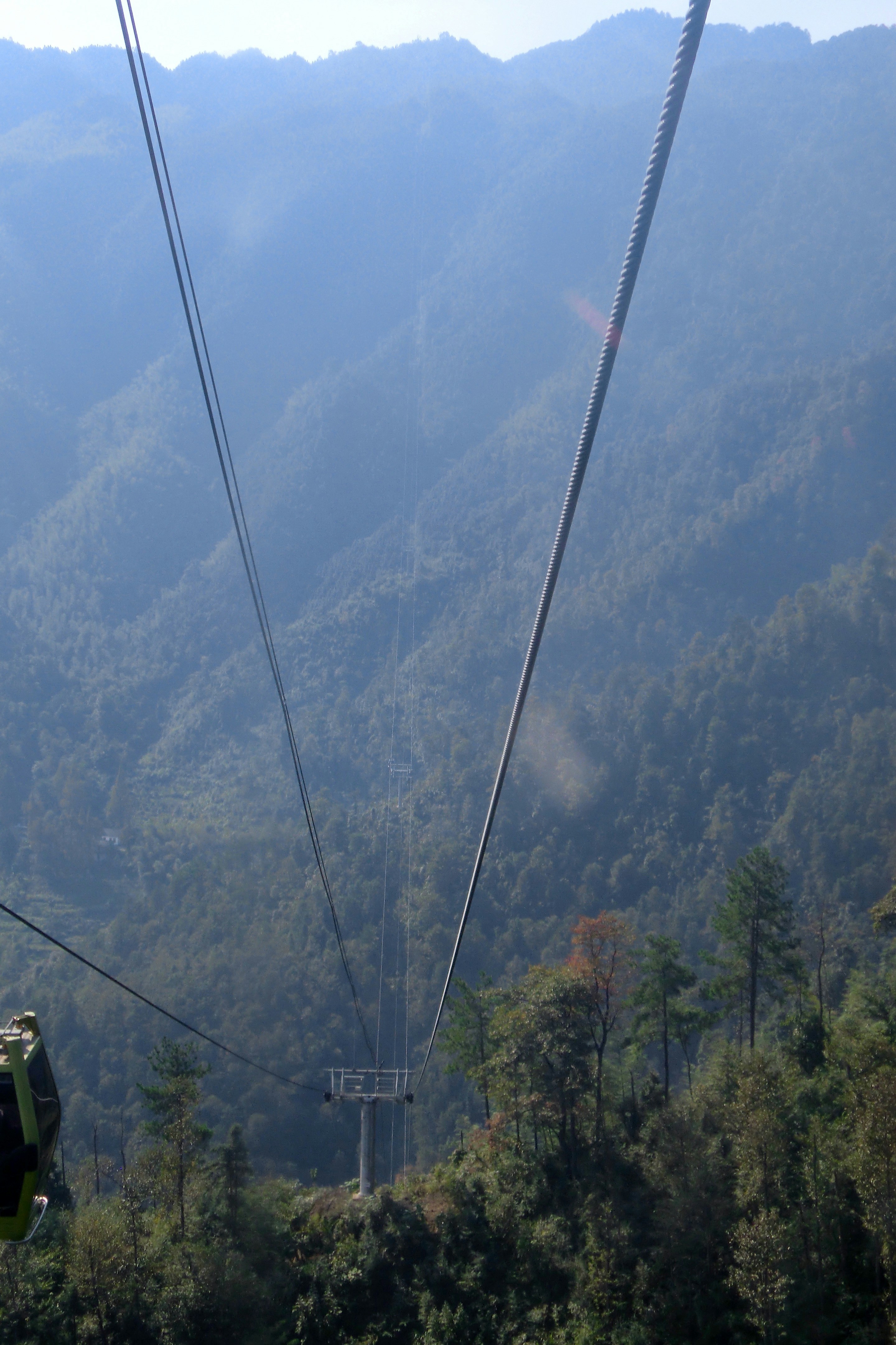 Cable car lines stretch across a misty alpine valley, with a steel tower rising amid forested slopes.