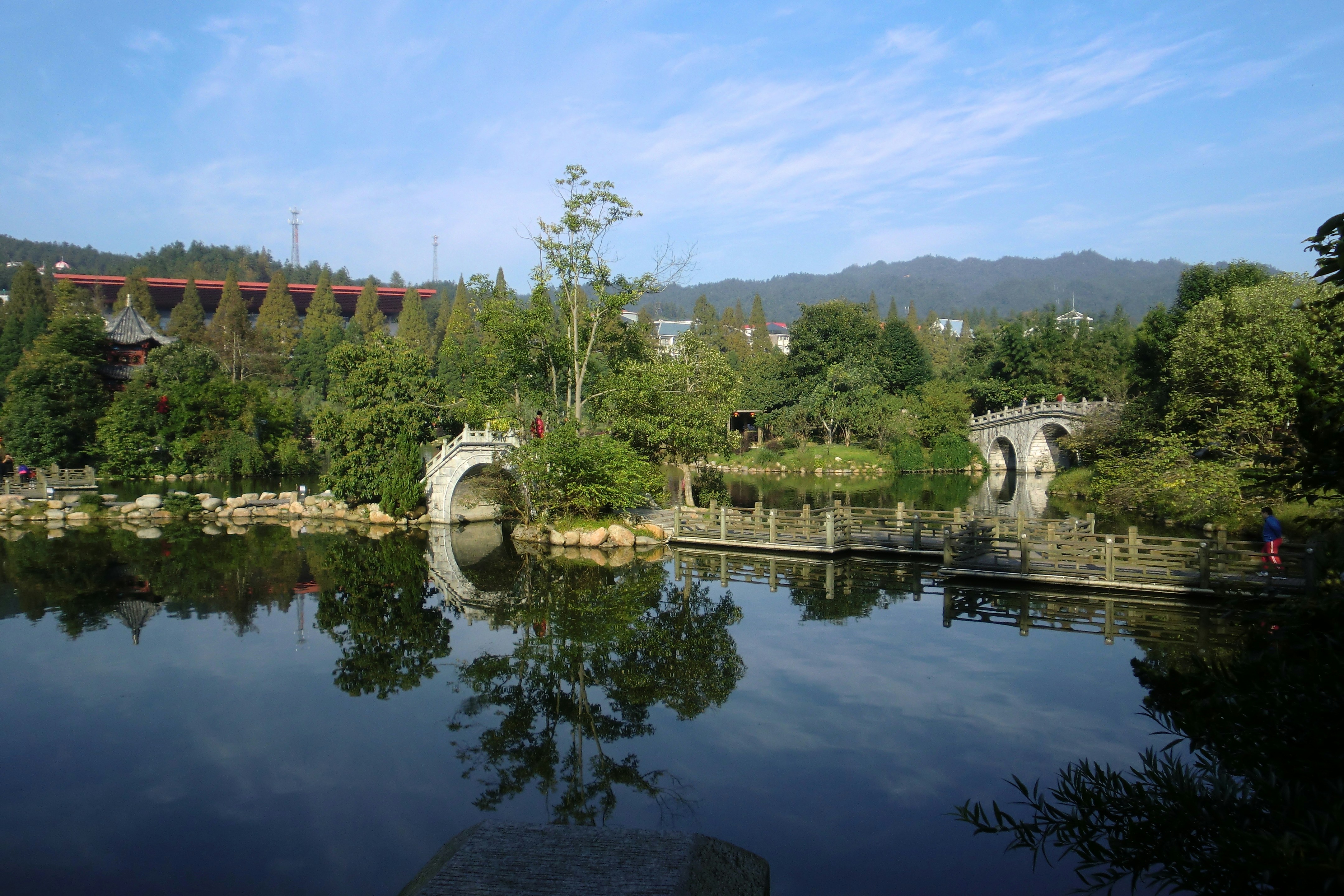 A tranquil garden scene featuring arched stone bridges and lush greenery, mirrored in calm waters under a clear blue sky.