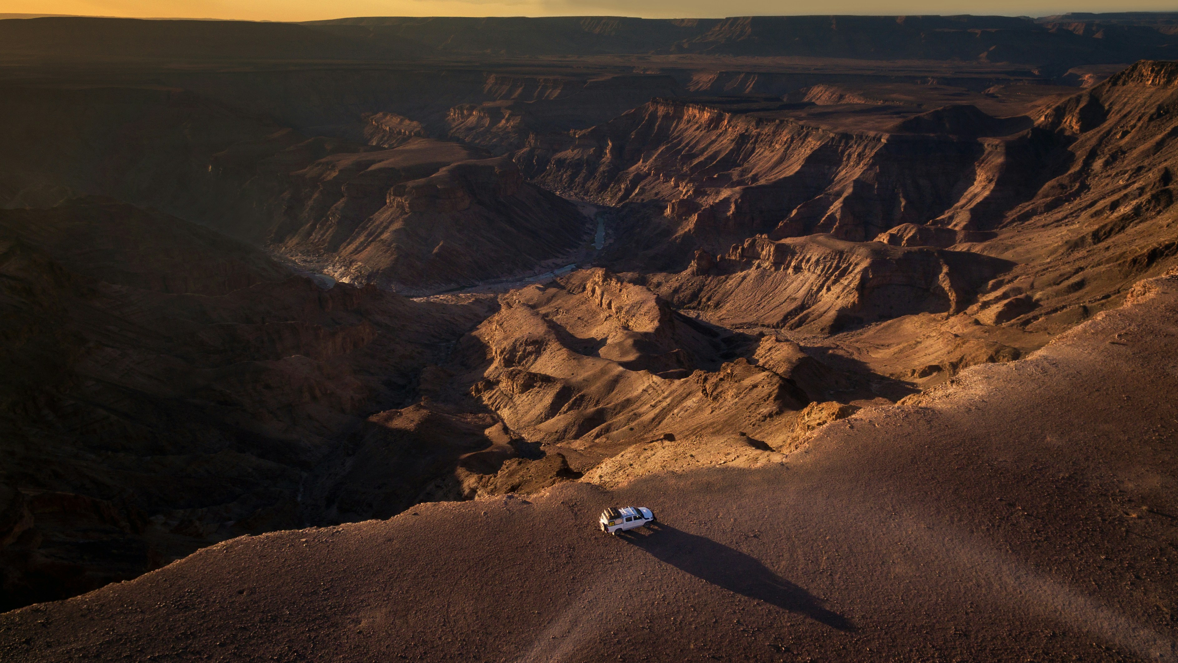 A small white drone hovers near the ridgeline above a deep canyon, casting a long shadow as the sun low on the horizon highlights the rugged rock formations.