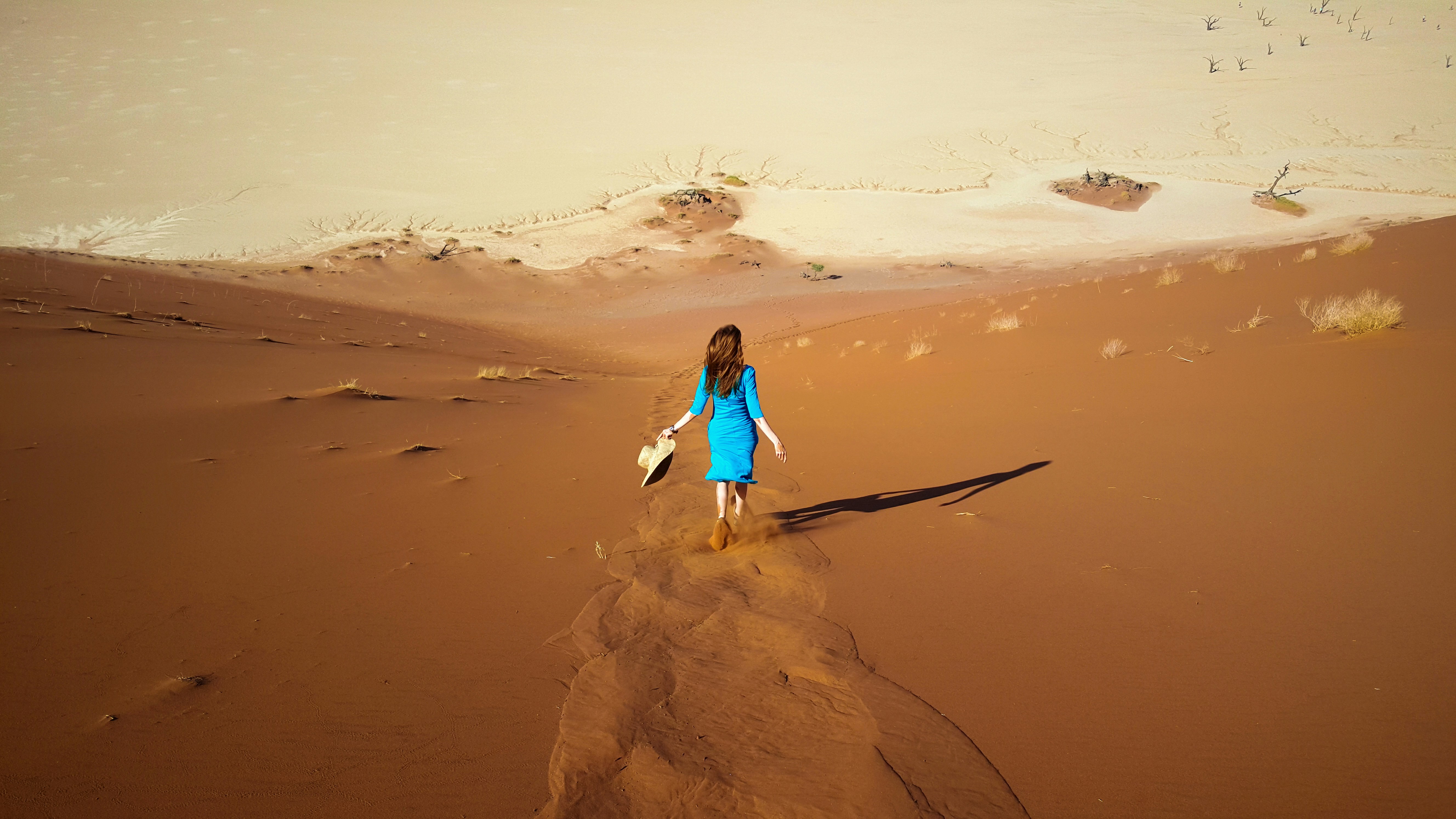 Photograph of a girl in a bright blue dress walking along sunlit sand toward the water, leaving footprints behind.
