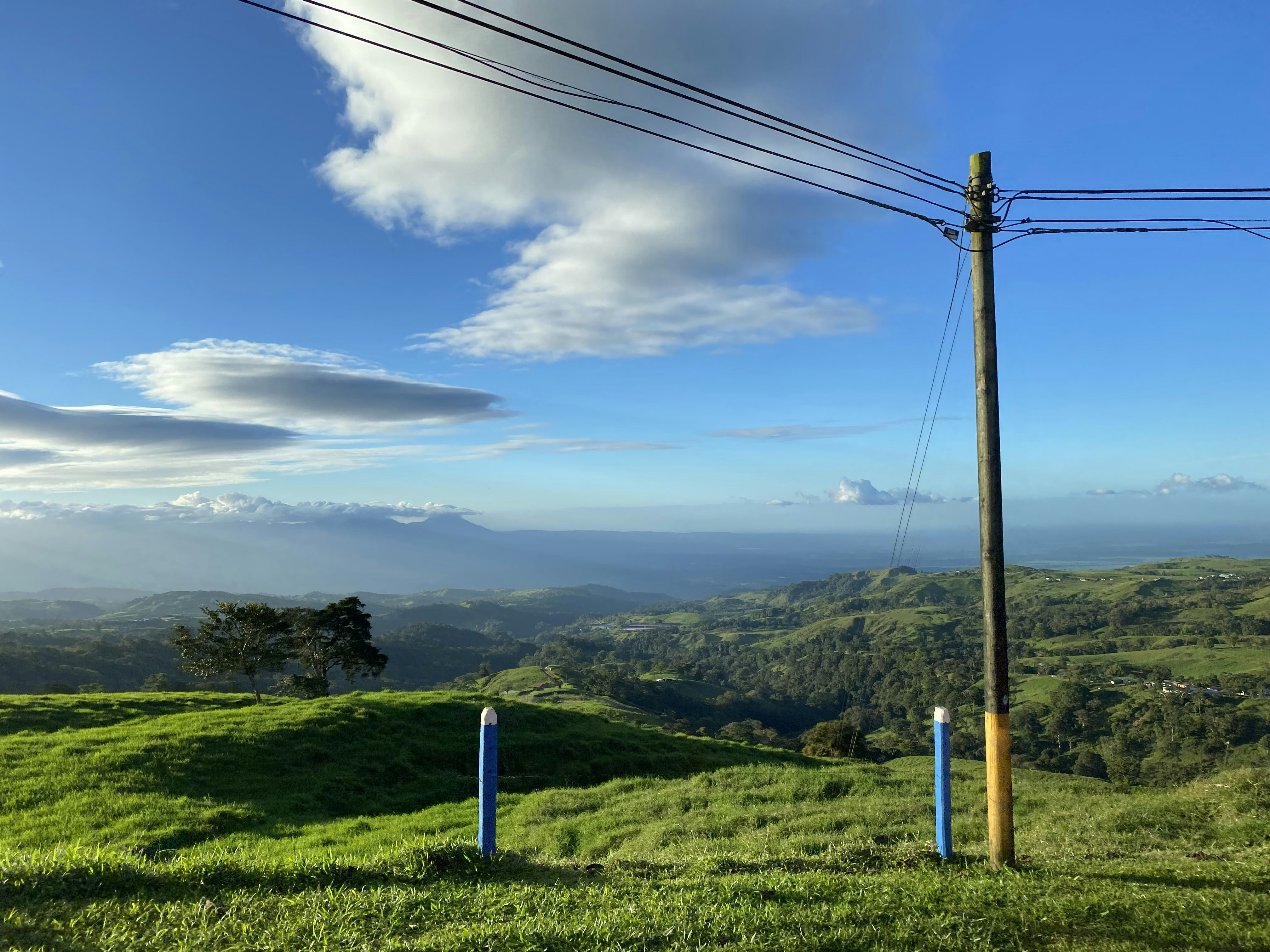 Expansive green hills rolling towards the ocean under a clear blue sky, framed by utility poles and wispy clouds.