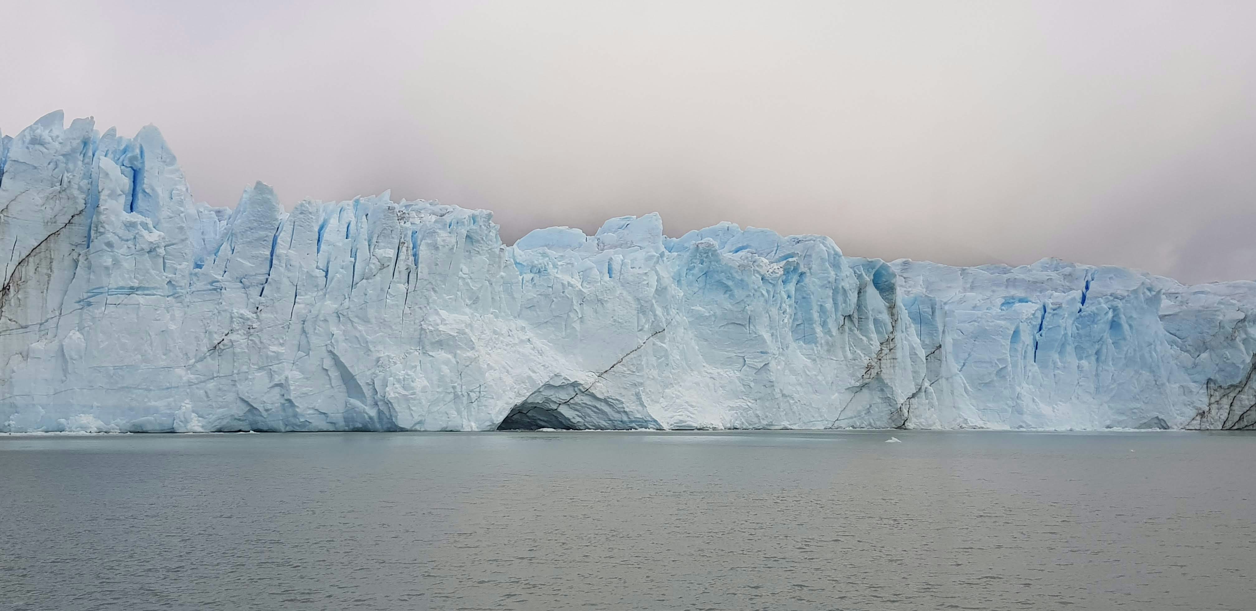 Expansive glacier with rugged ice formations stretching across a tranquil body of water under an overcast sky.
