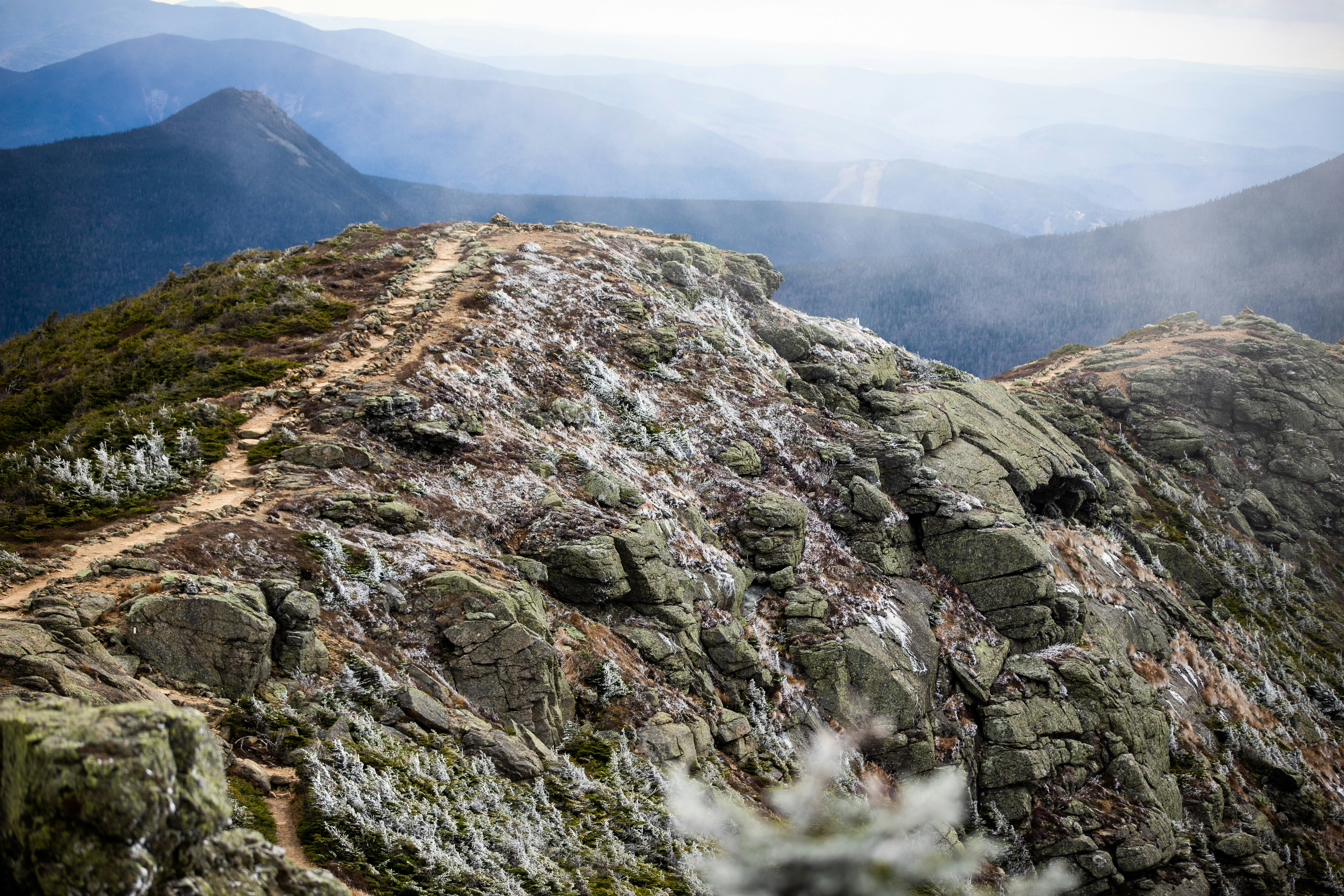 Winding trail across a rocky ridge, surrounded by misty mountains and sparse vegetation. The rugged terrain showcases nature's raw beauty.