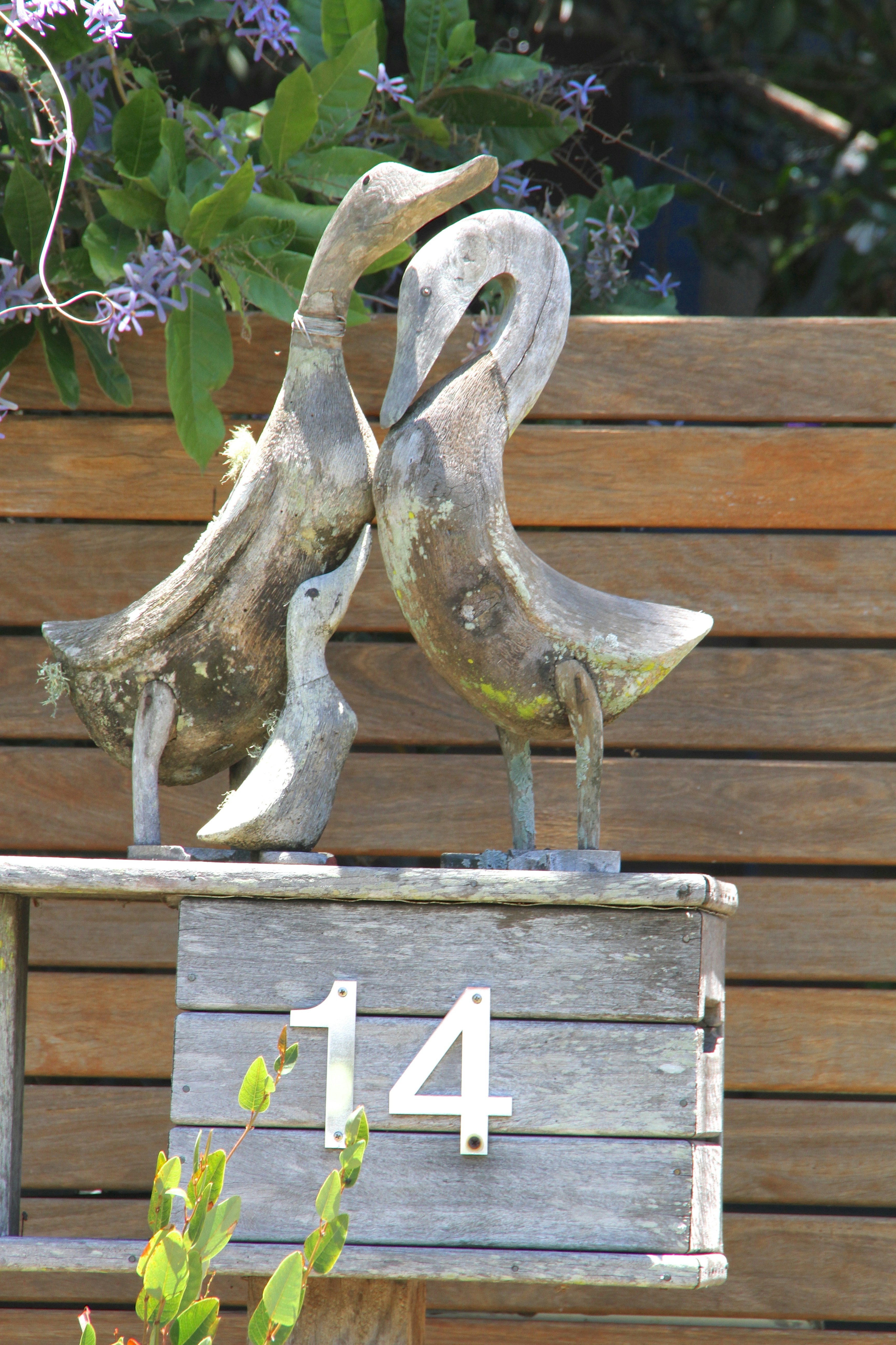 Bronze duck sculptures nestled on a wooden mailbox, surrounded by lush greenery and purple flowers.