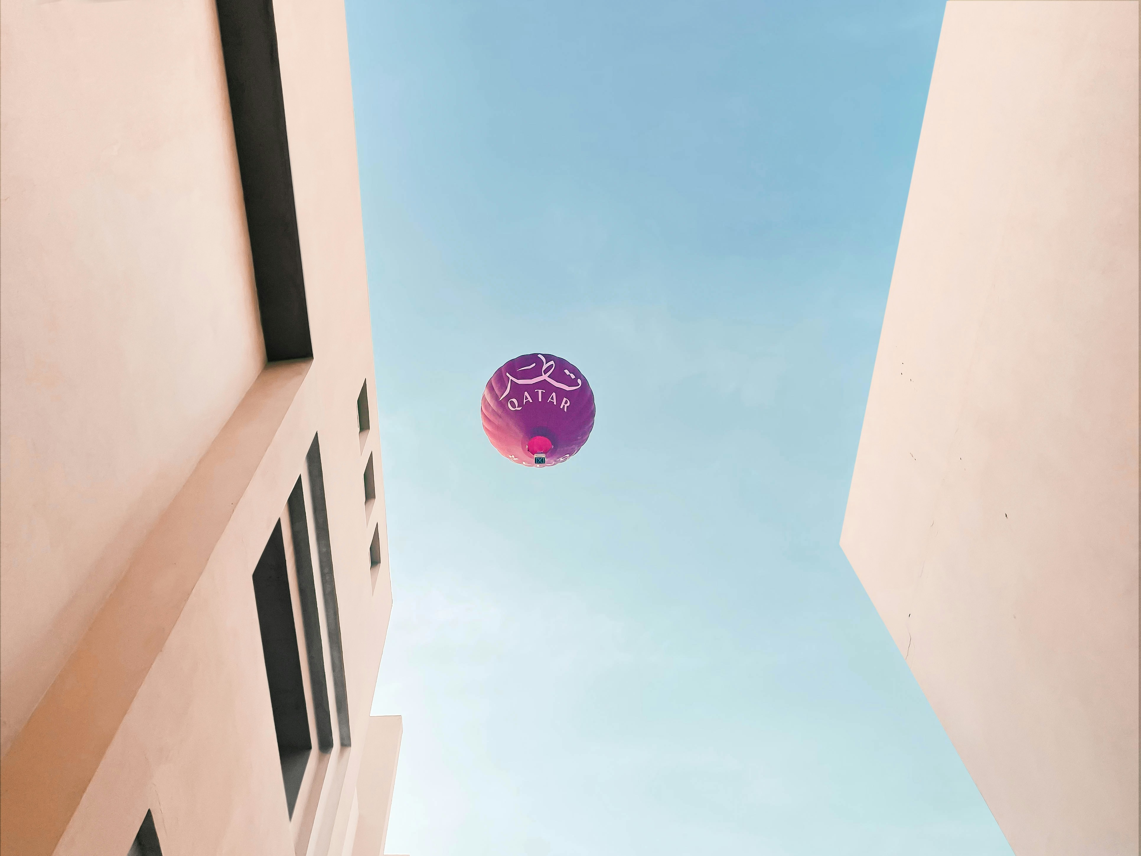 Colorful hot air balloon floating above modern architecture, framed by angular buildings against a clear blue sky.