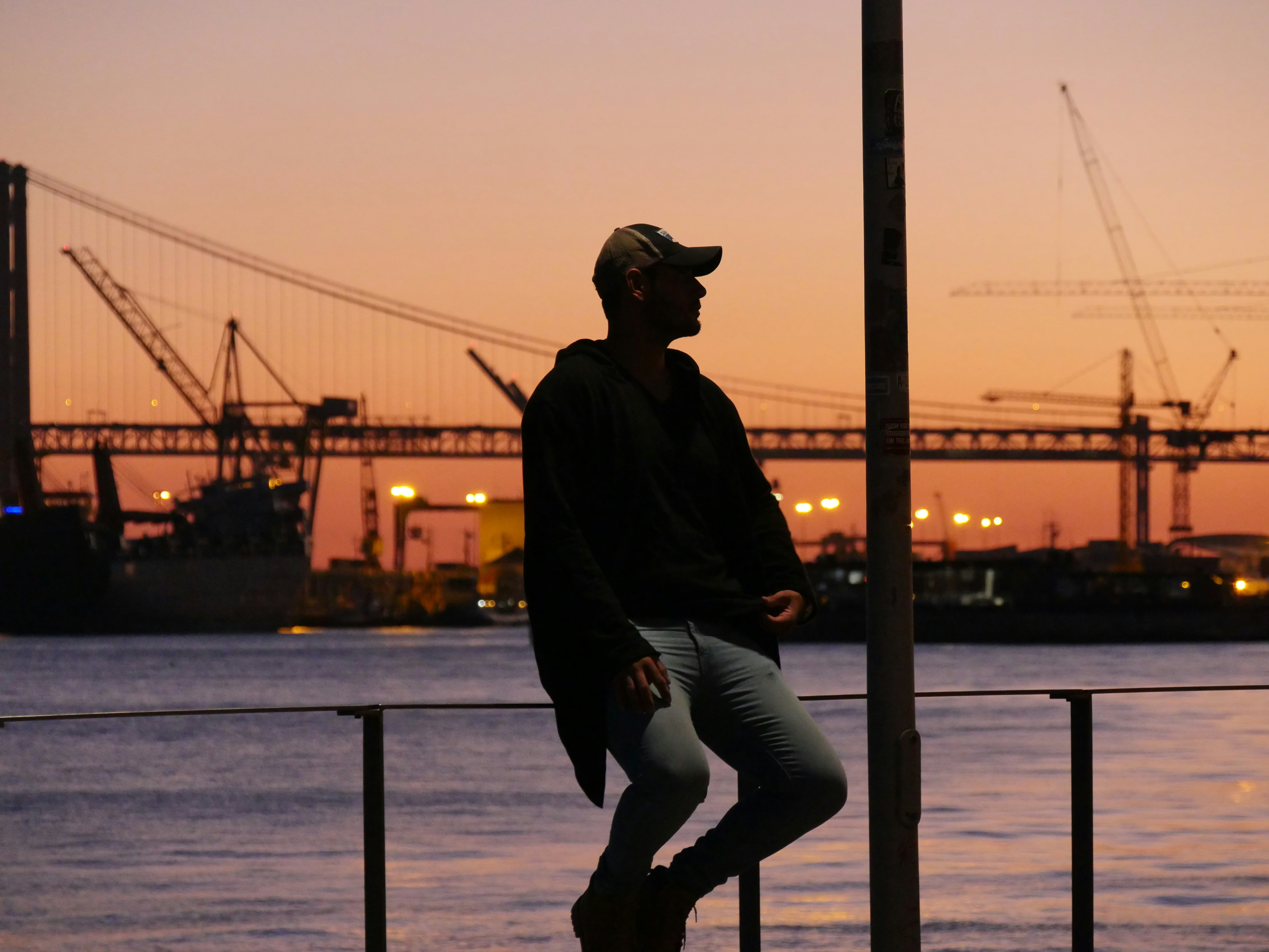 Dusk Silhouette by the Harbor: A lone figure framed by cranes and glow