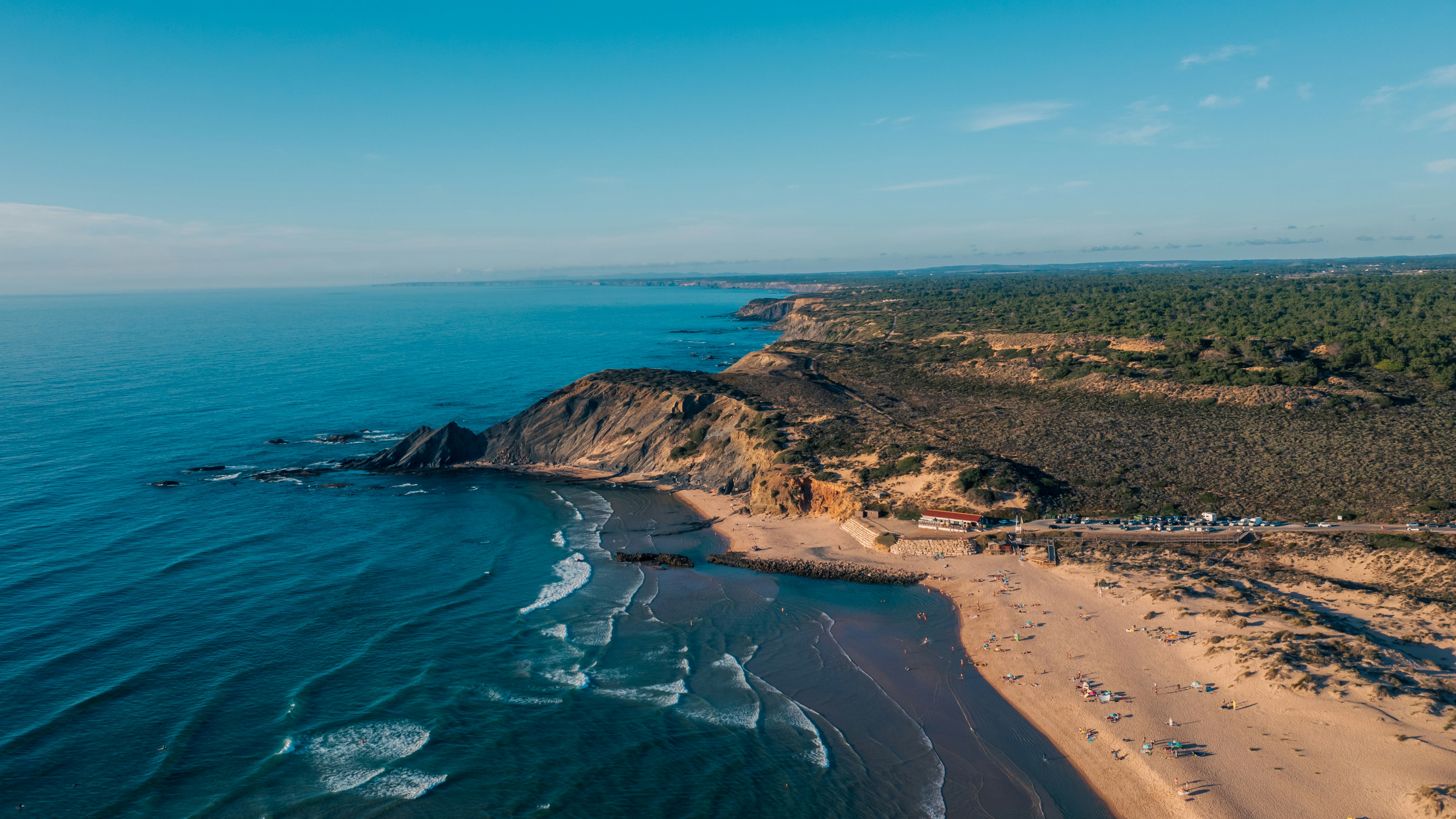Aerial view of a picturesque beach with gentle waves lapping at the shore, bordered by rugged cliffs and lush greenery in the background.