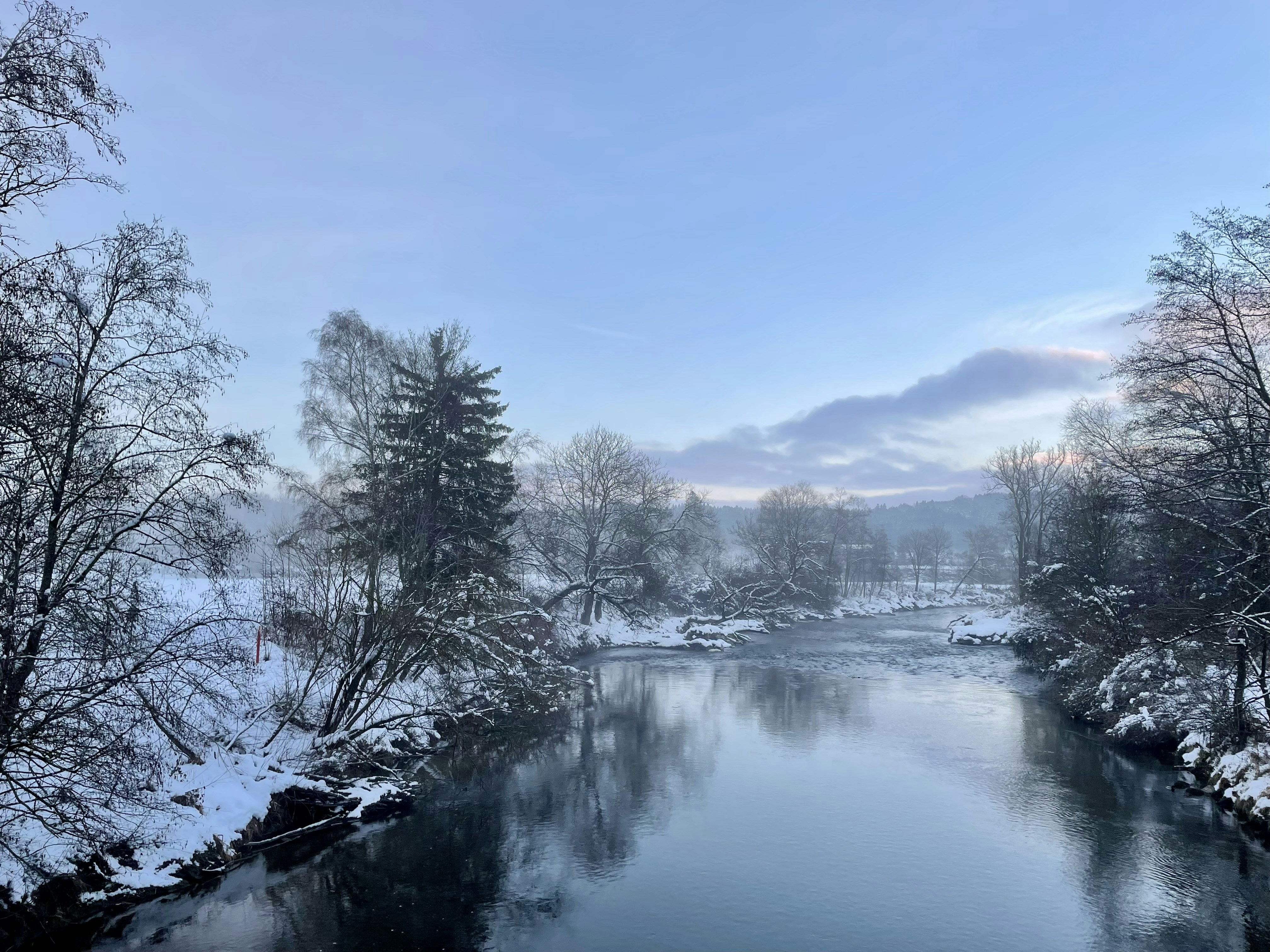 Tranquil river flowing through a snowy landscape, framed by frosted trees under a soft blue sky.