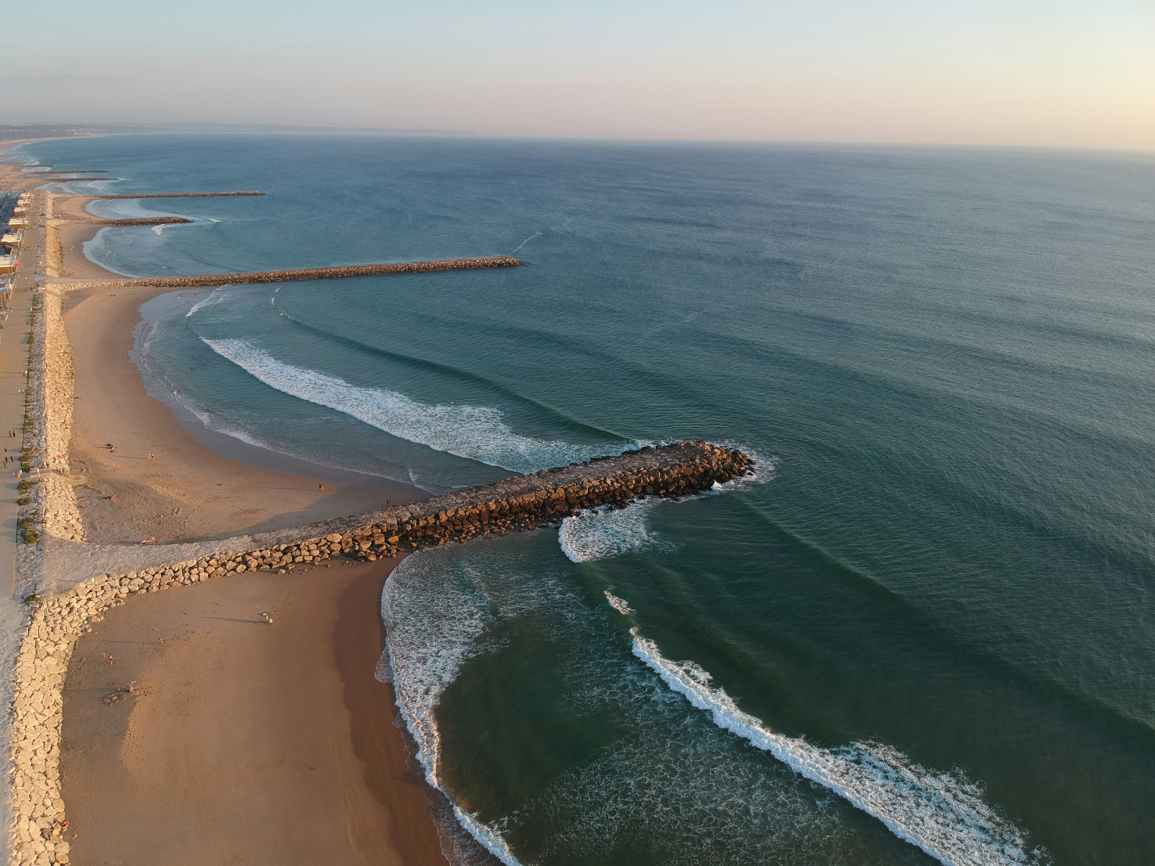 Aerial view of a sandy beach and calm ocean waves meeting a jetty at dawn.